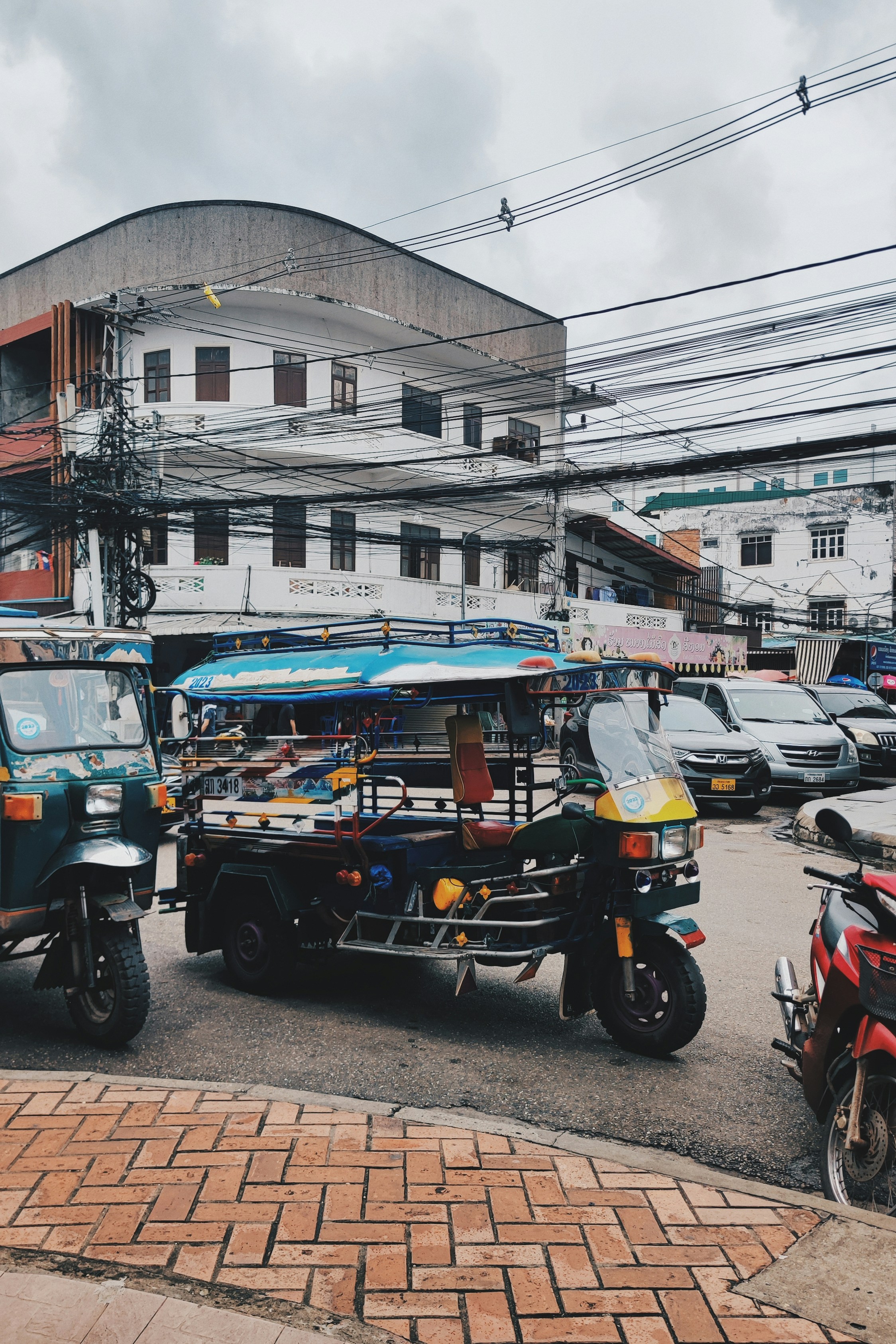 Colorful tuk-tuks parked amidst bustling streets and tangled power lines in a vibrant urban setting.