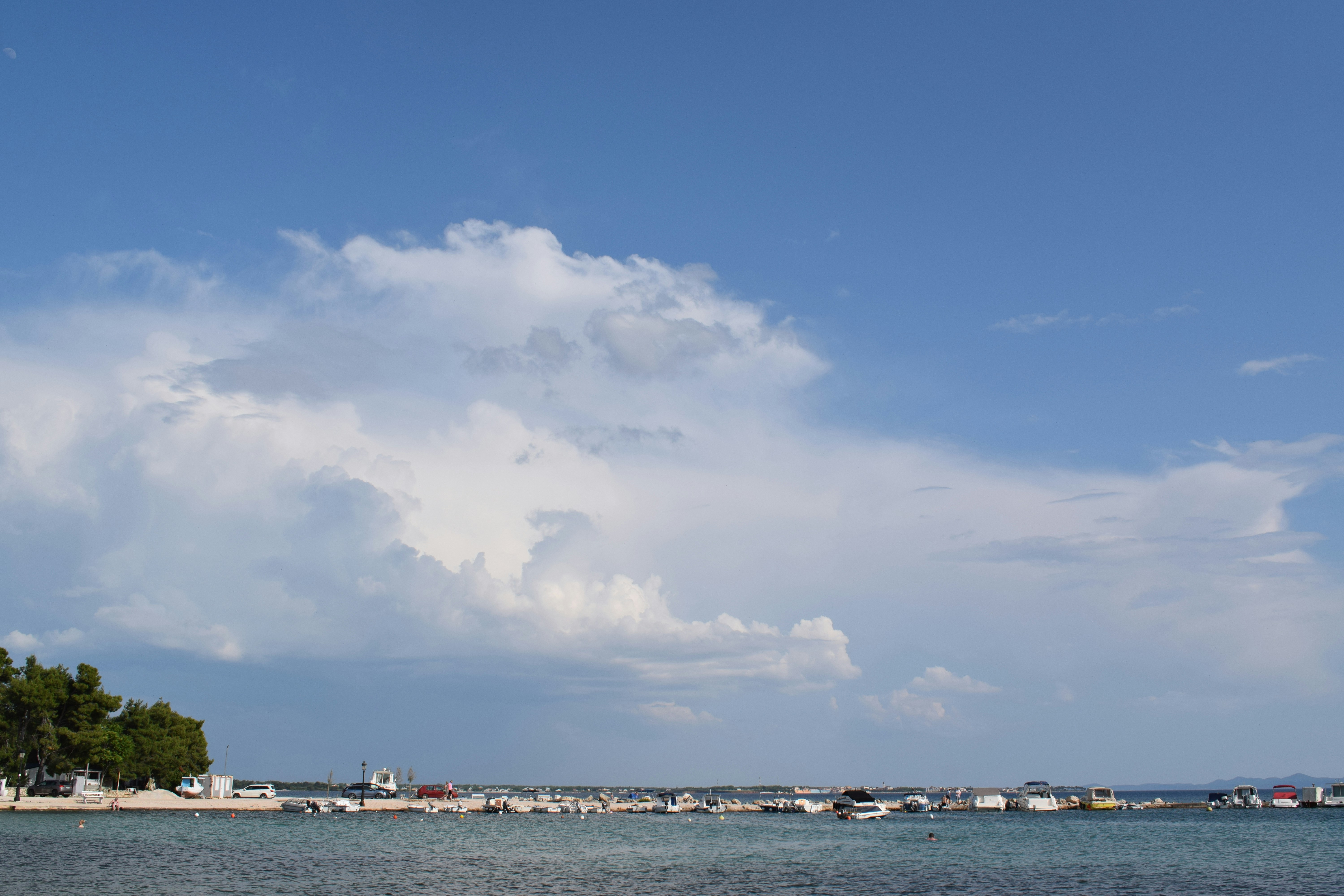 A tranquil harbor scene with boats anchored along the shoreline, framed by lush greenery and a vast, cloud-studded sky. 
