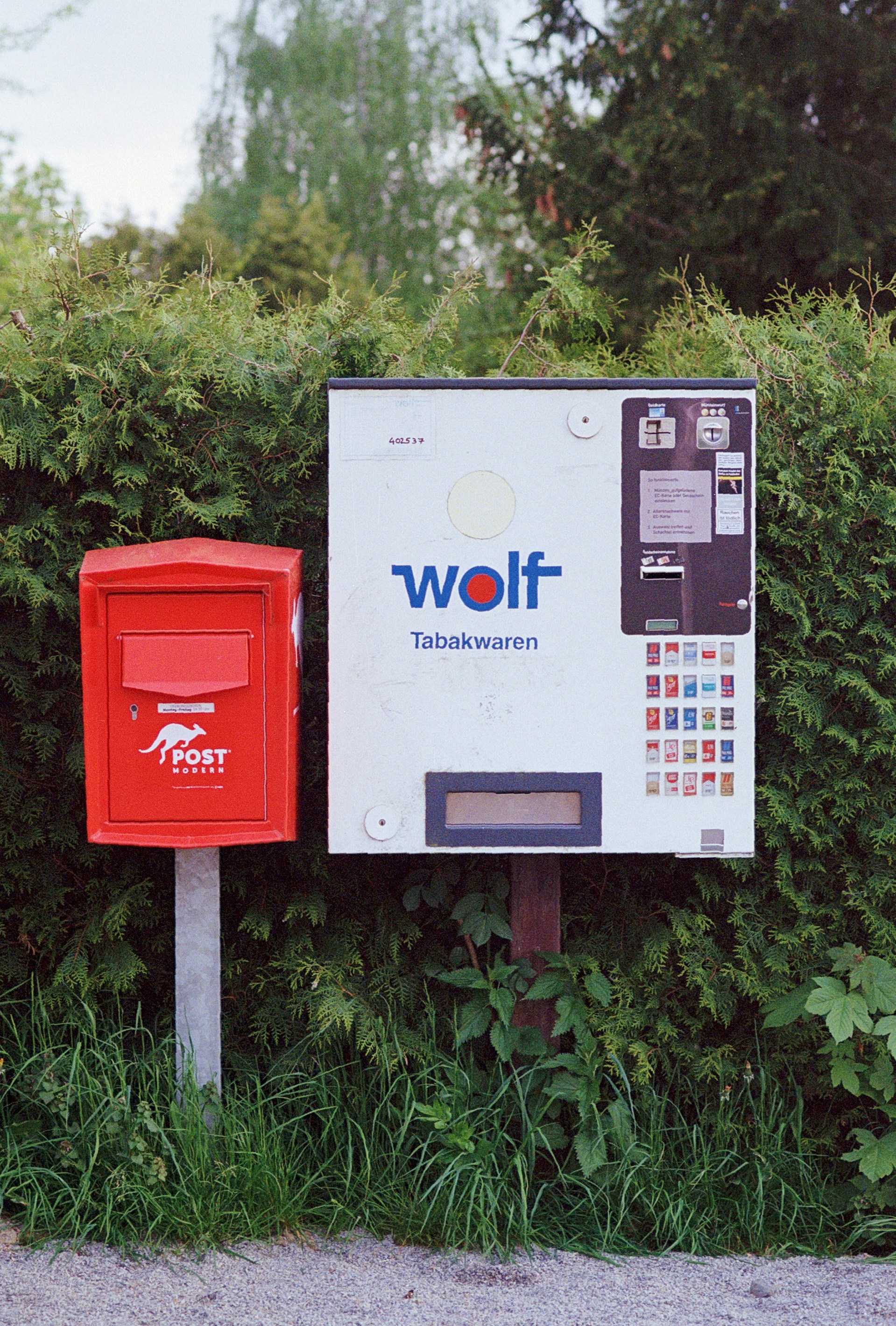 Red Post Box and Wolf Tabakwaren Cabinet Nestled Against a Hedge