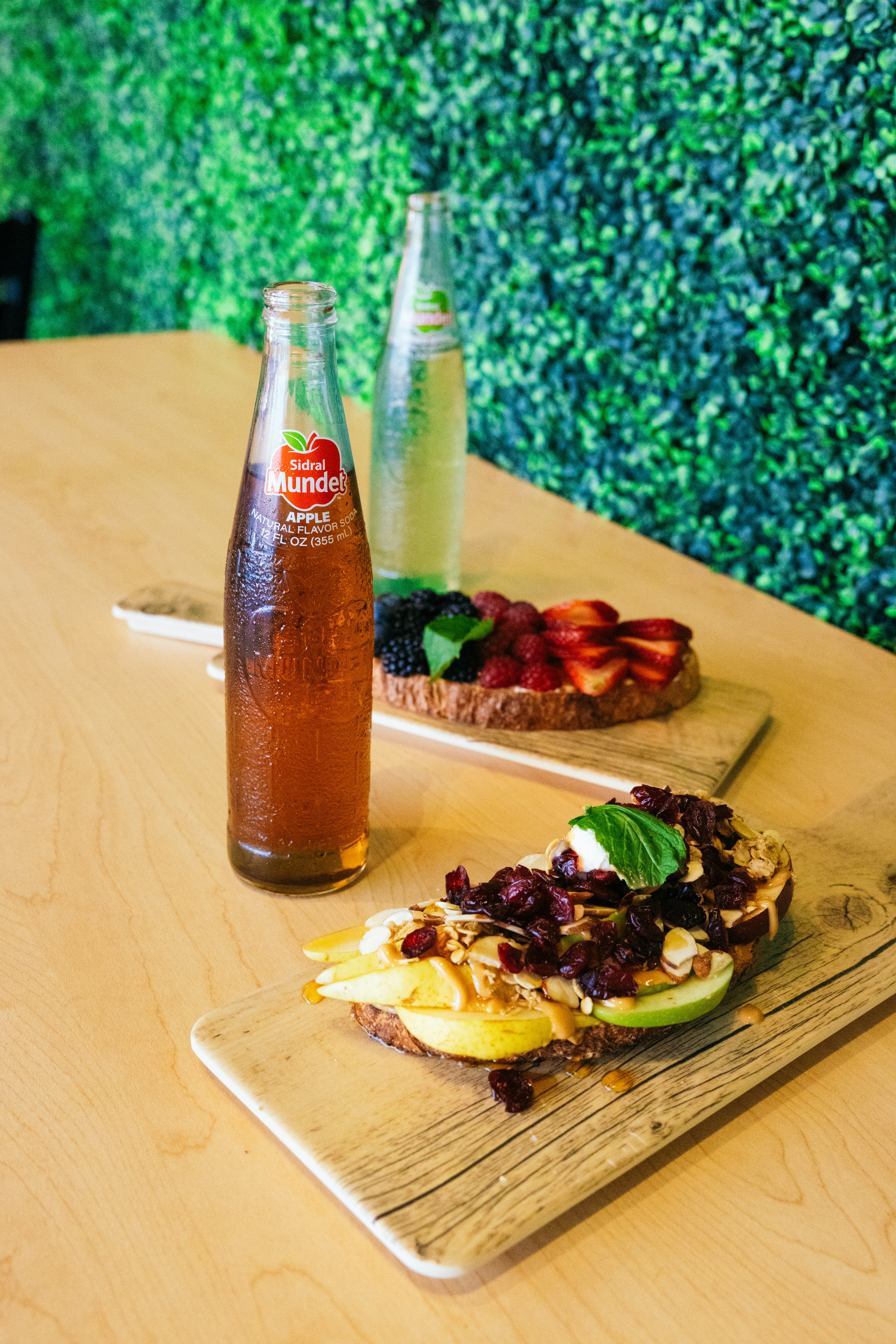 Photograph of an amber apple soda bottle in focus on a wooden table with a fruit-topped toast in the foreground, set against a green leafy backdrop.