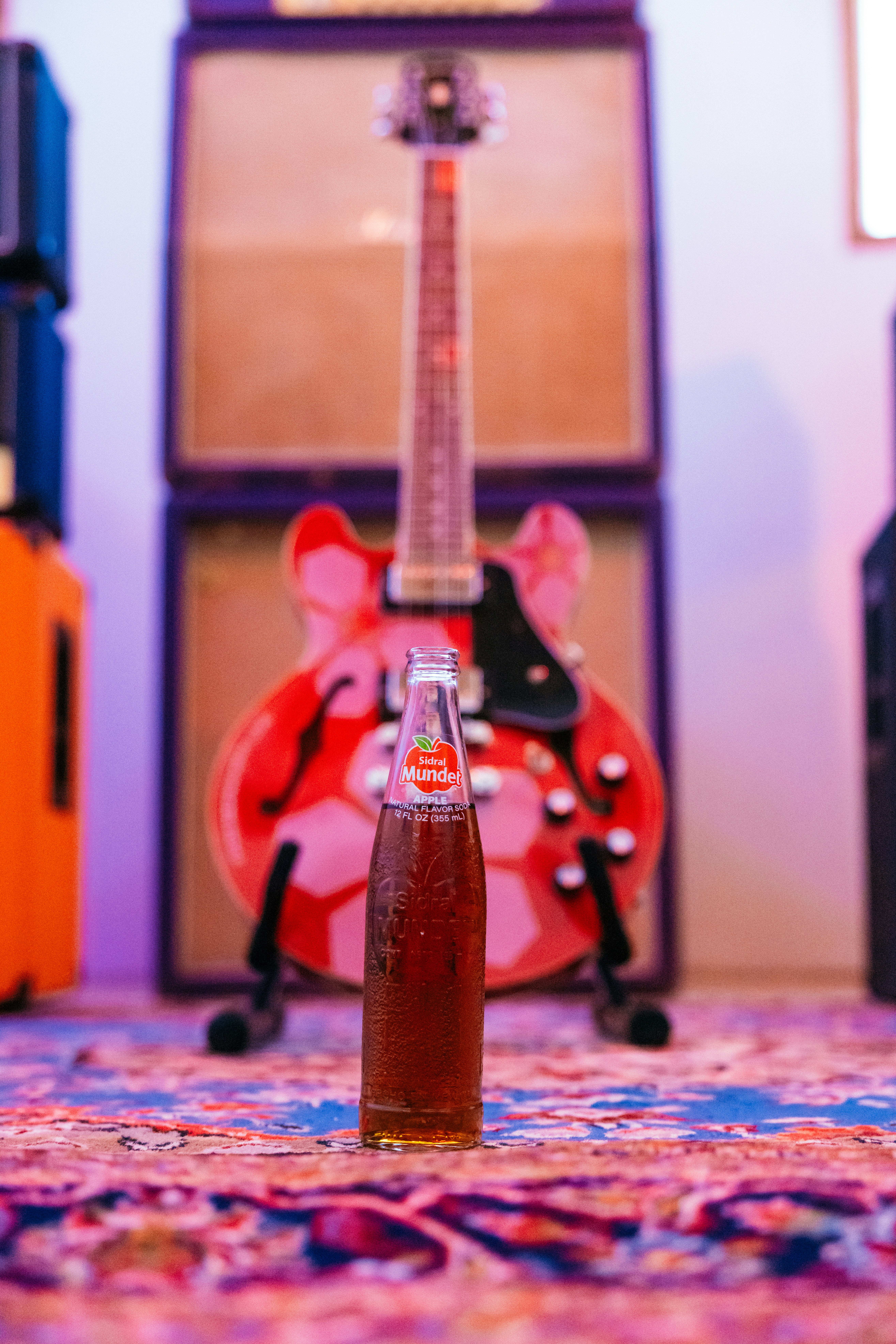 A bottle sits centered on a patterned rug with a pink guitar and amplifiers blurred in the background.