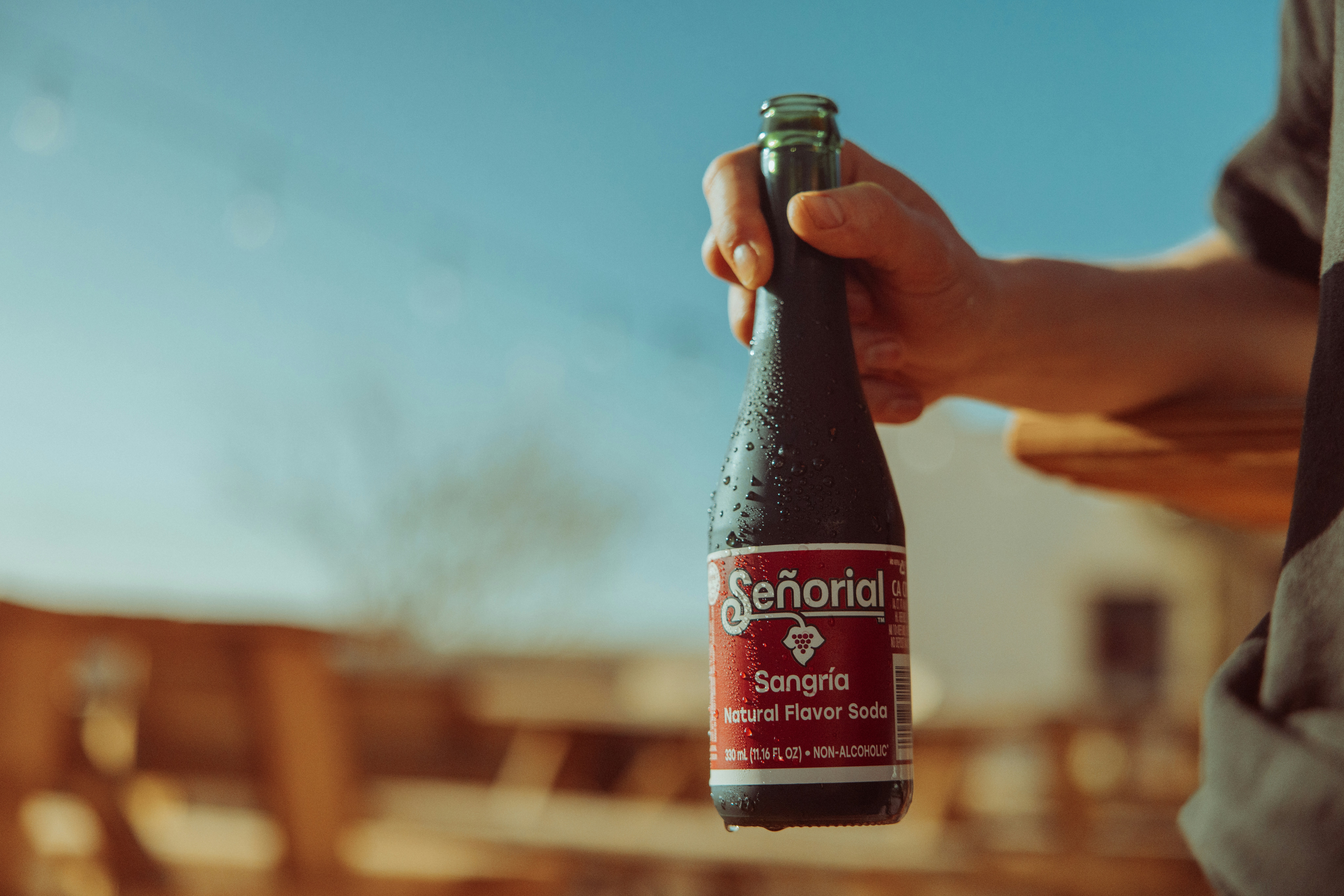 Close-up of a Senorial Sangria bottle held by a hand on a sunlit patio, with a blurred wooden deck and blue sky in the background.