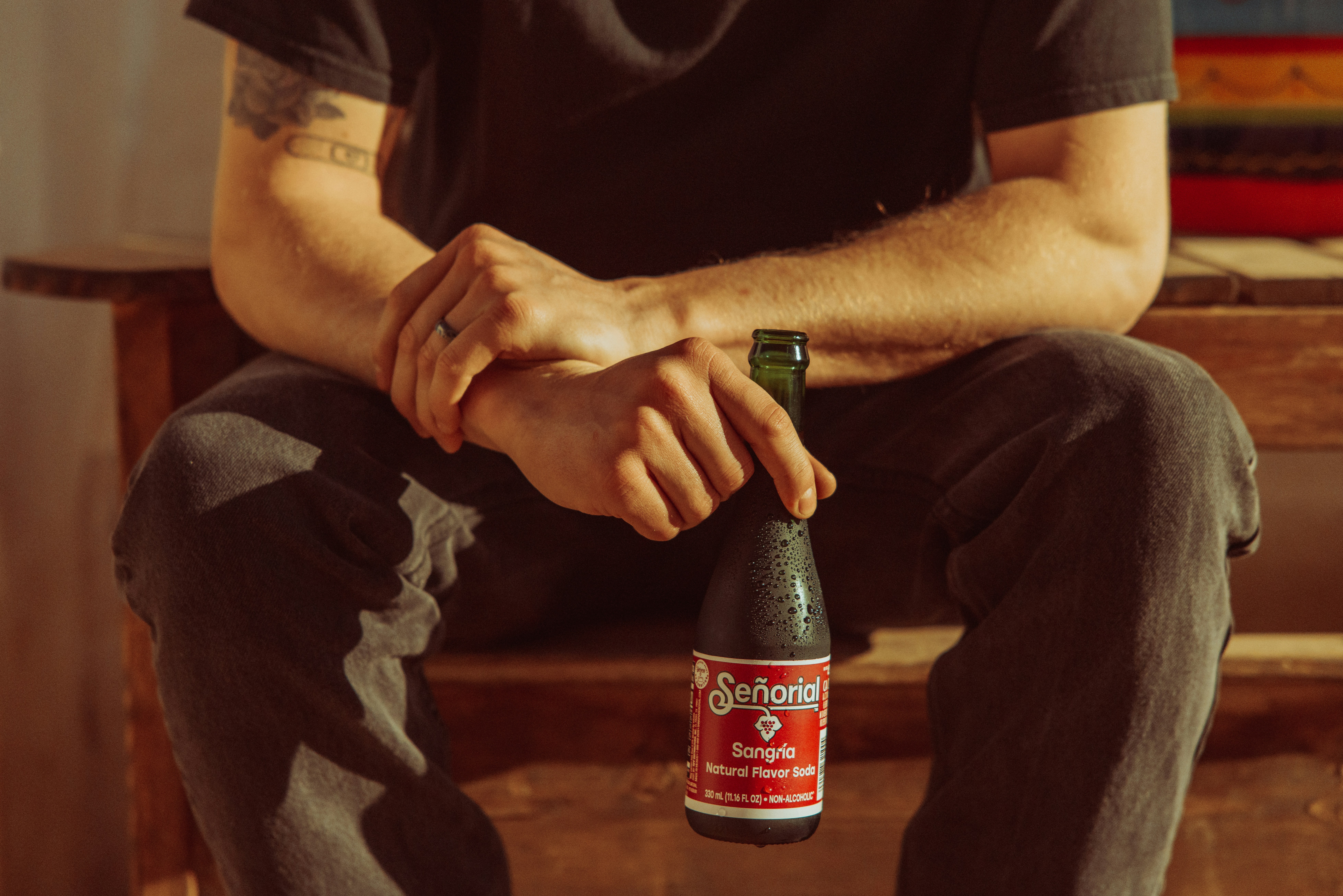 A man sits on wooden steps, hands resting on a beer bottle held between his fingers, under warm ambient light. This candid photograph focuses on a simple, everyday moment.