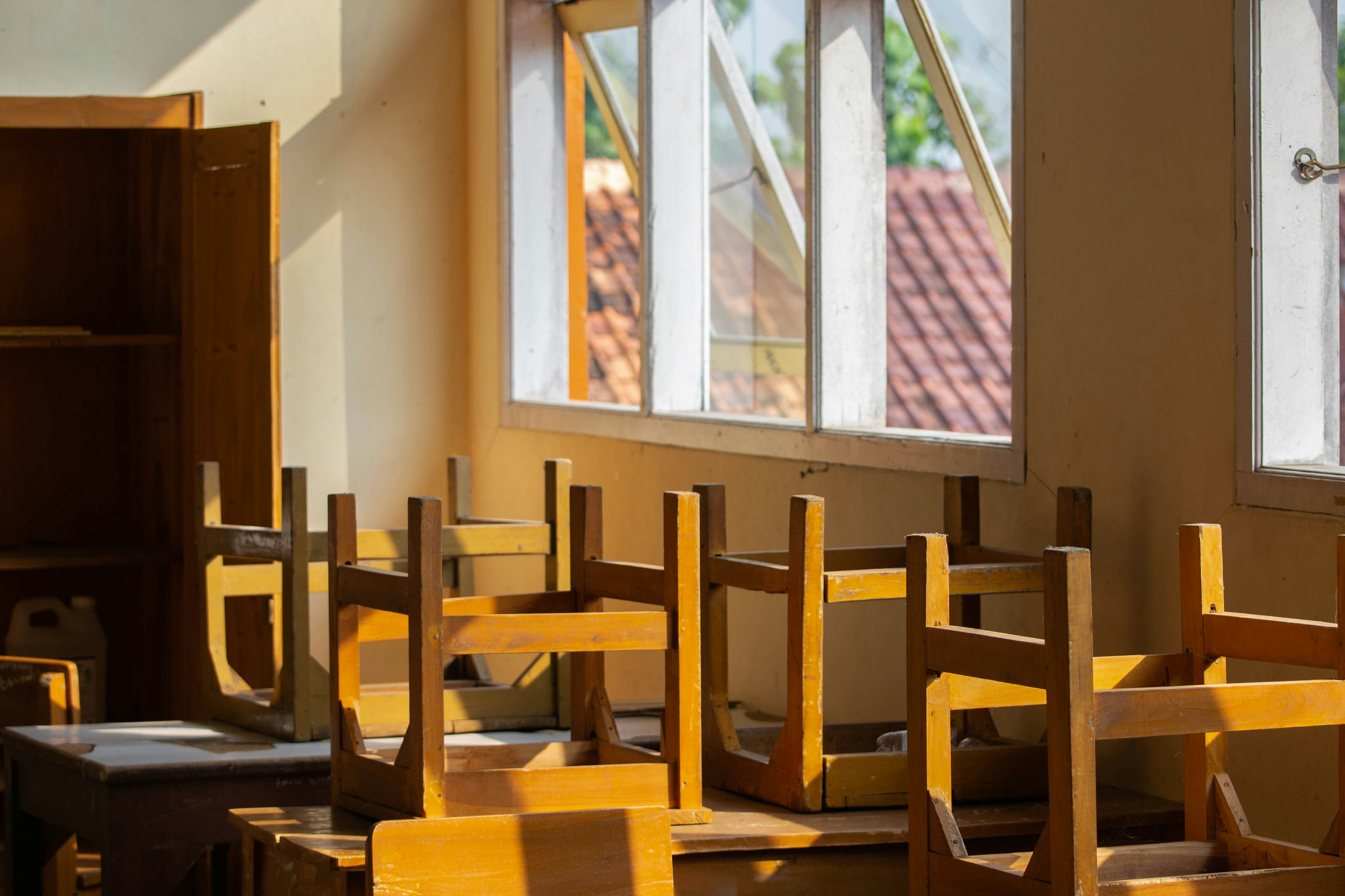 Upside-down chairs on desks in a sunlit classroom with large windows casting shadows.