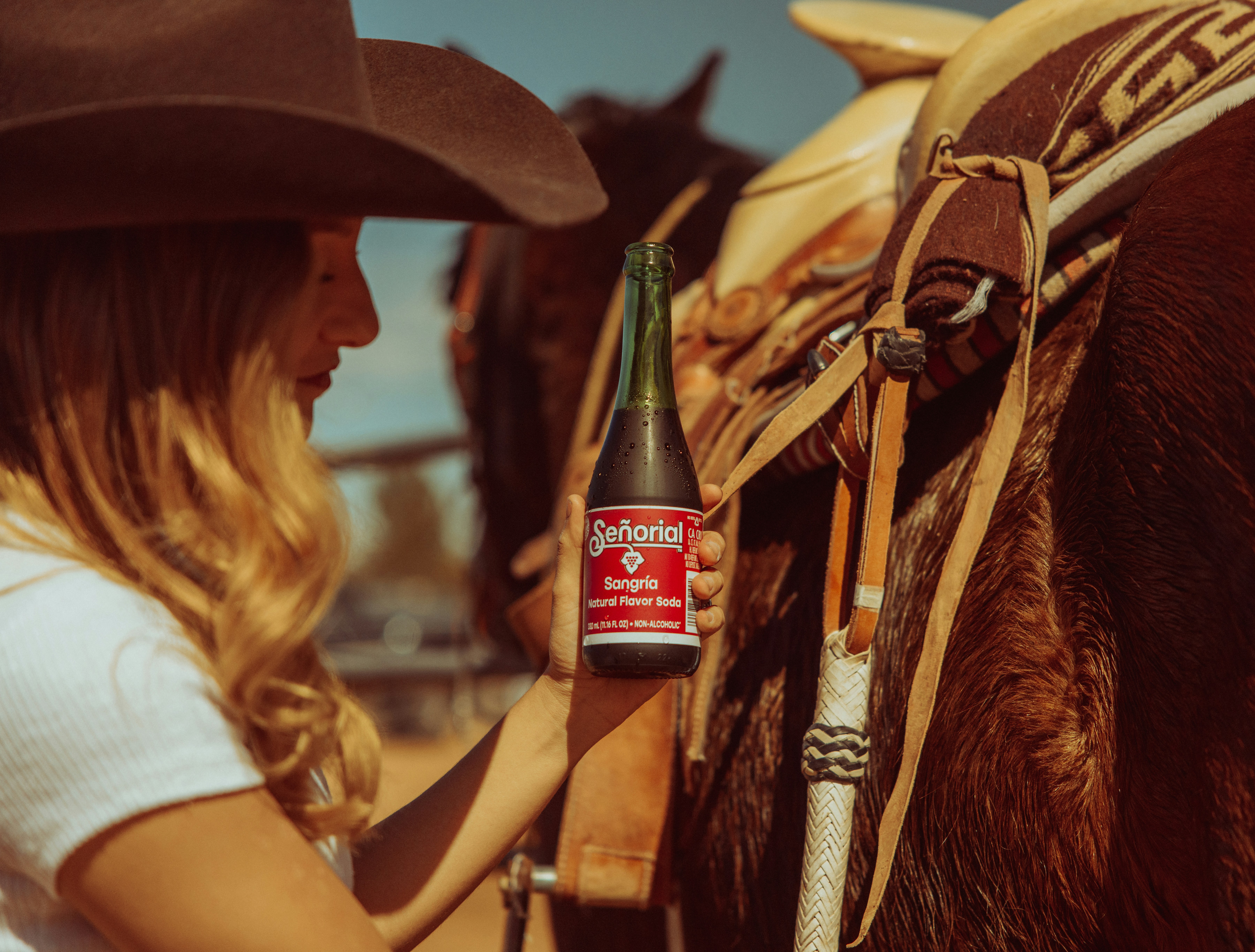 A young woman in a cowboy hat holds a bottle of Señorial Sangría next to a saddle on a horse, embodying the spirit of Western culture.