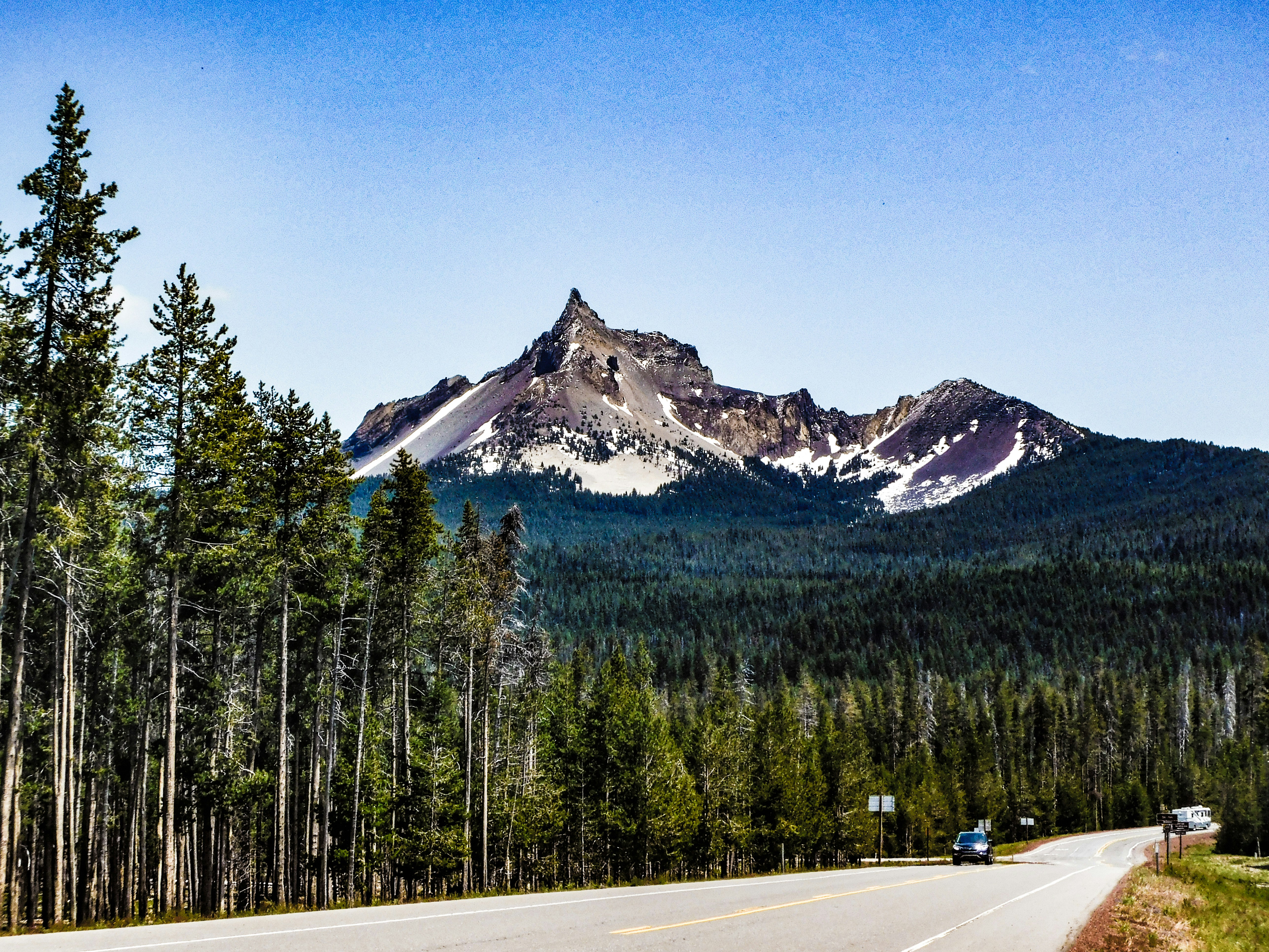 Mountain landscape photograph featuring a snow-dusted peak, dense pine forest, and a curving highway.
