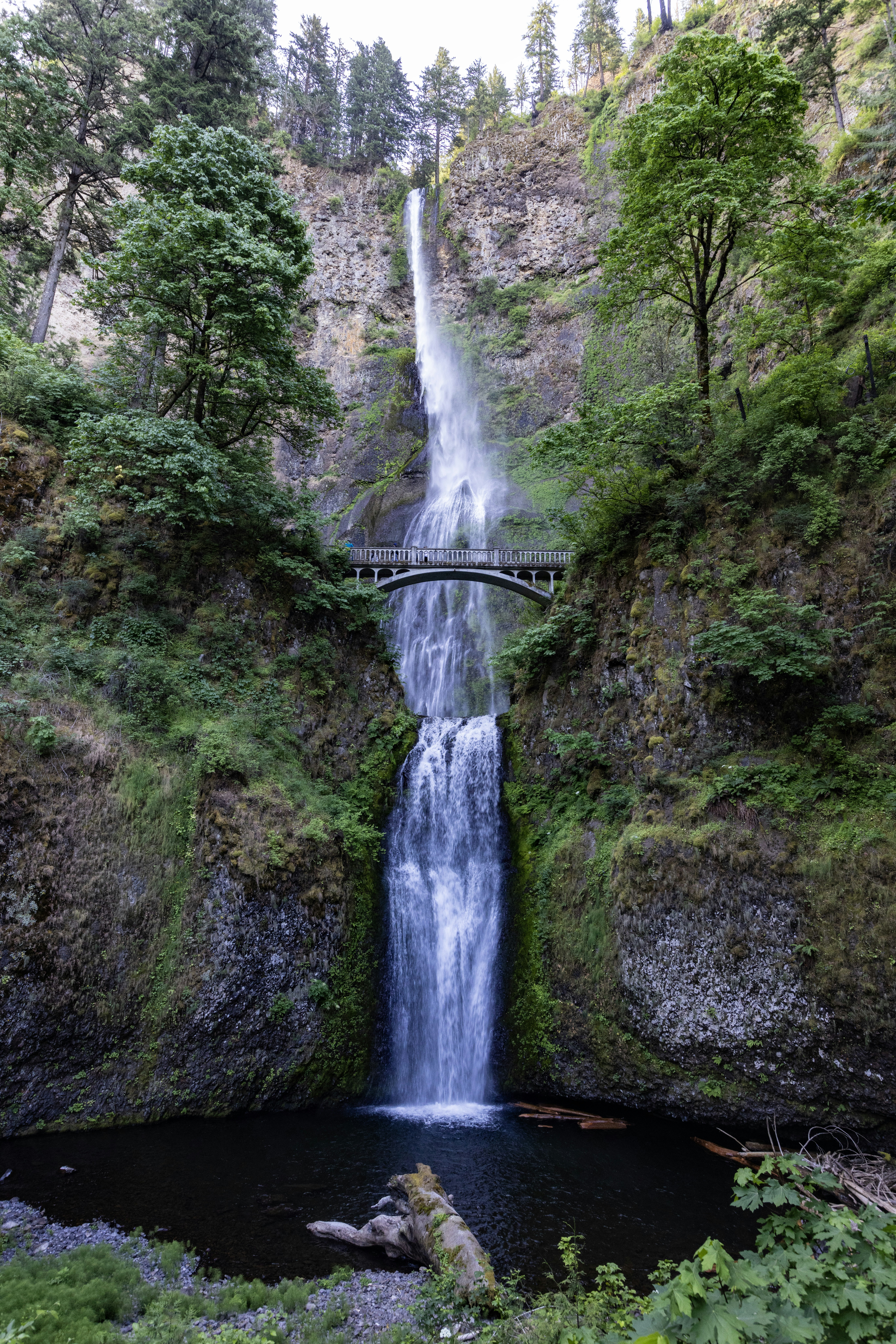 Majestic waterfall cascading down rocky cliffs, framed by lush greenery and a quaint bridge above. The scene captures the harmony of water and landscape.