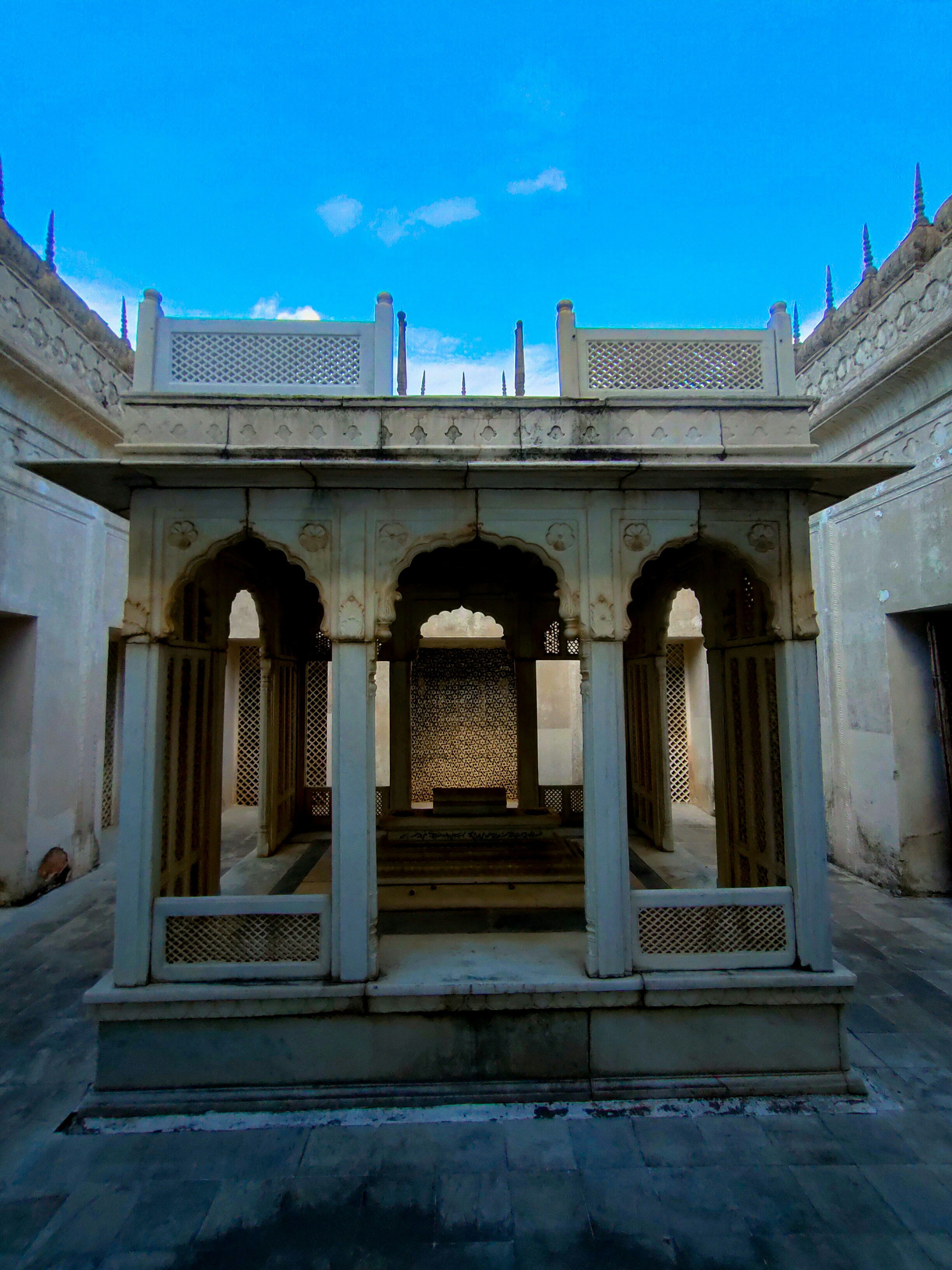 Intricate pavilion stands at the center of an ancient courtyard, framed by weathered walls and a vibrant blue sky above.