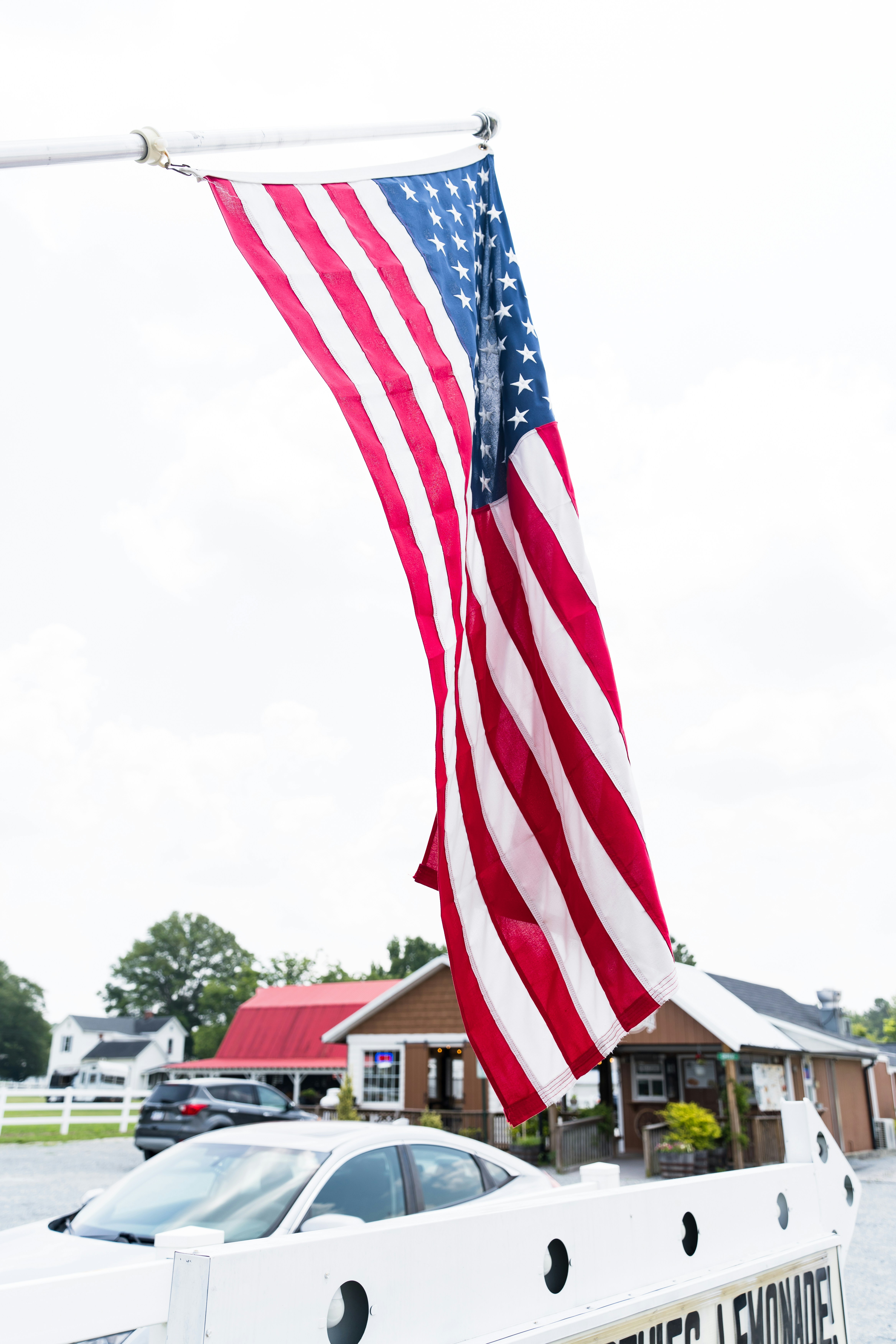 American flag billowing in the breeze, prominently displayed near a roadside lemonade stand. Bright colors contrast against a cloudy sky.