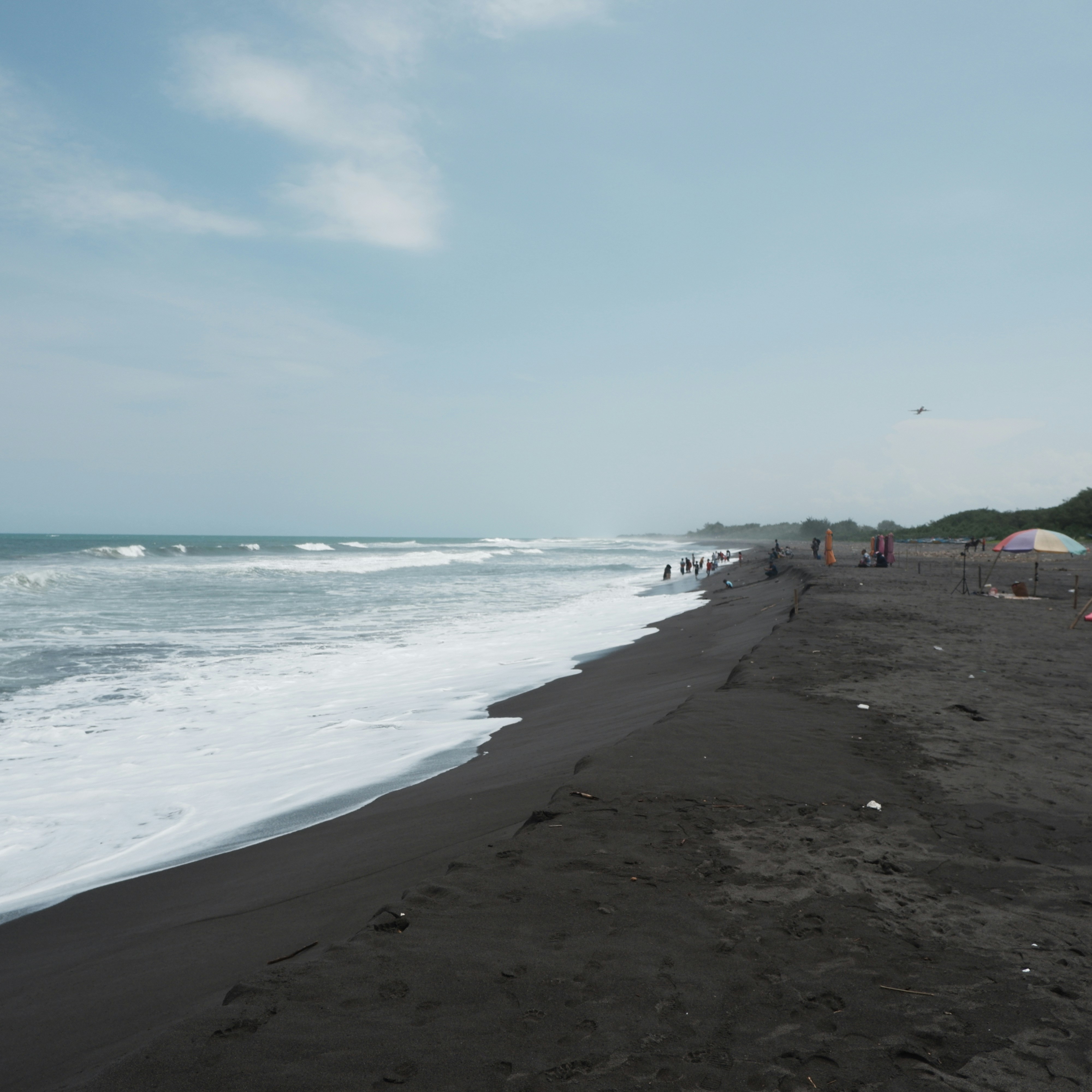 Waves gently lap at a black sand beach, with distant figures strolling along the shore under a clear sky.