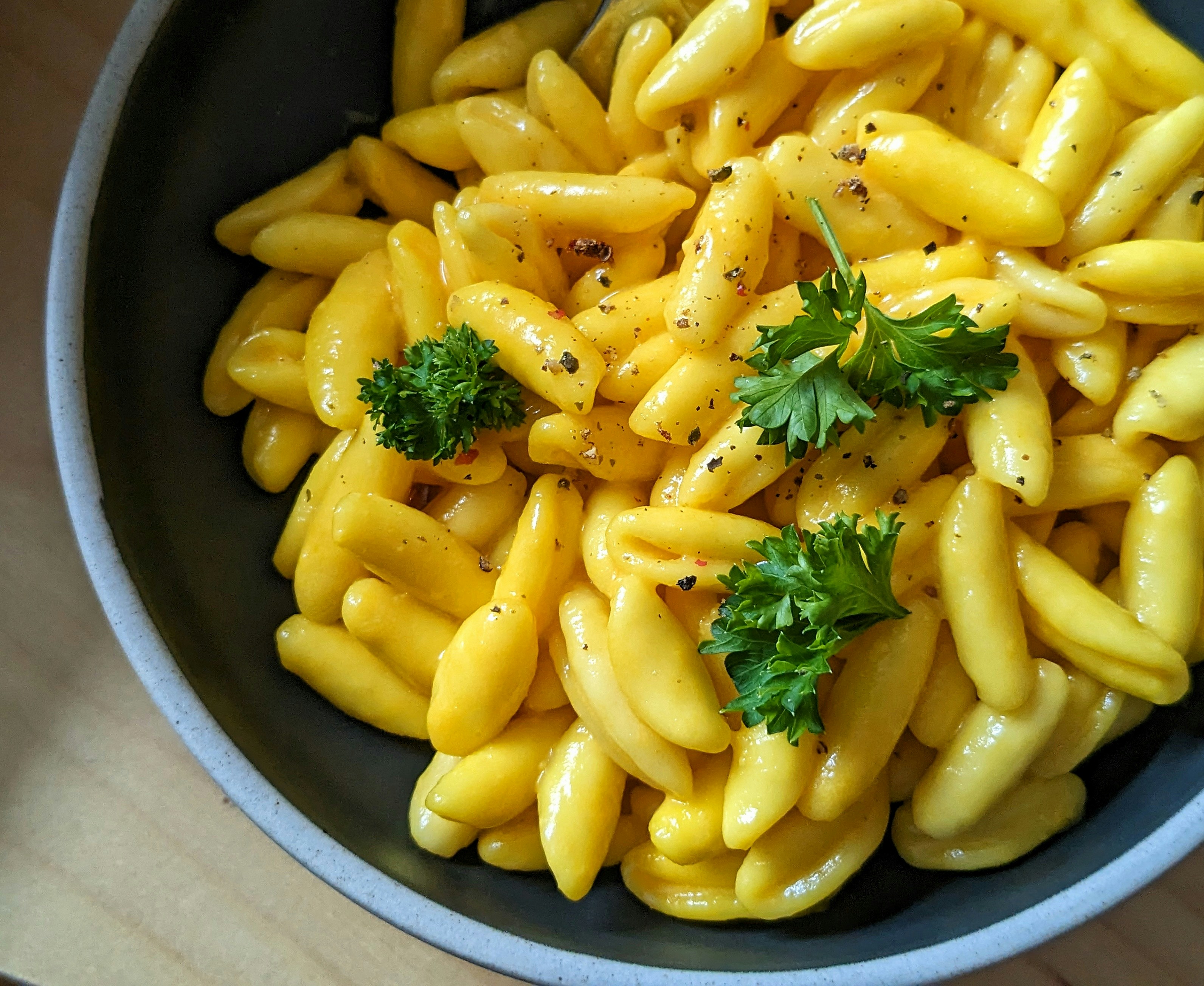 Close-up of golden penne pasta with a light glaze, garnished with fresh parsley in a matte ceramic bowl.
