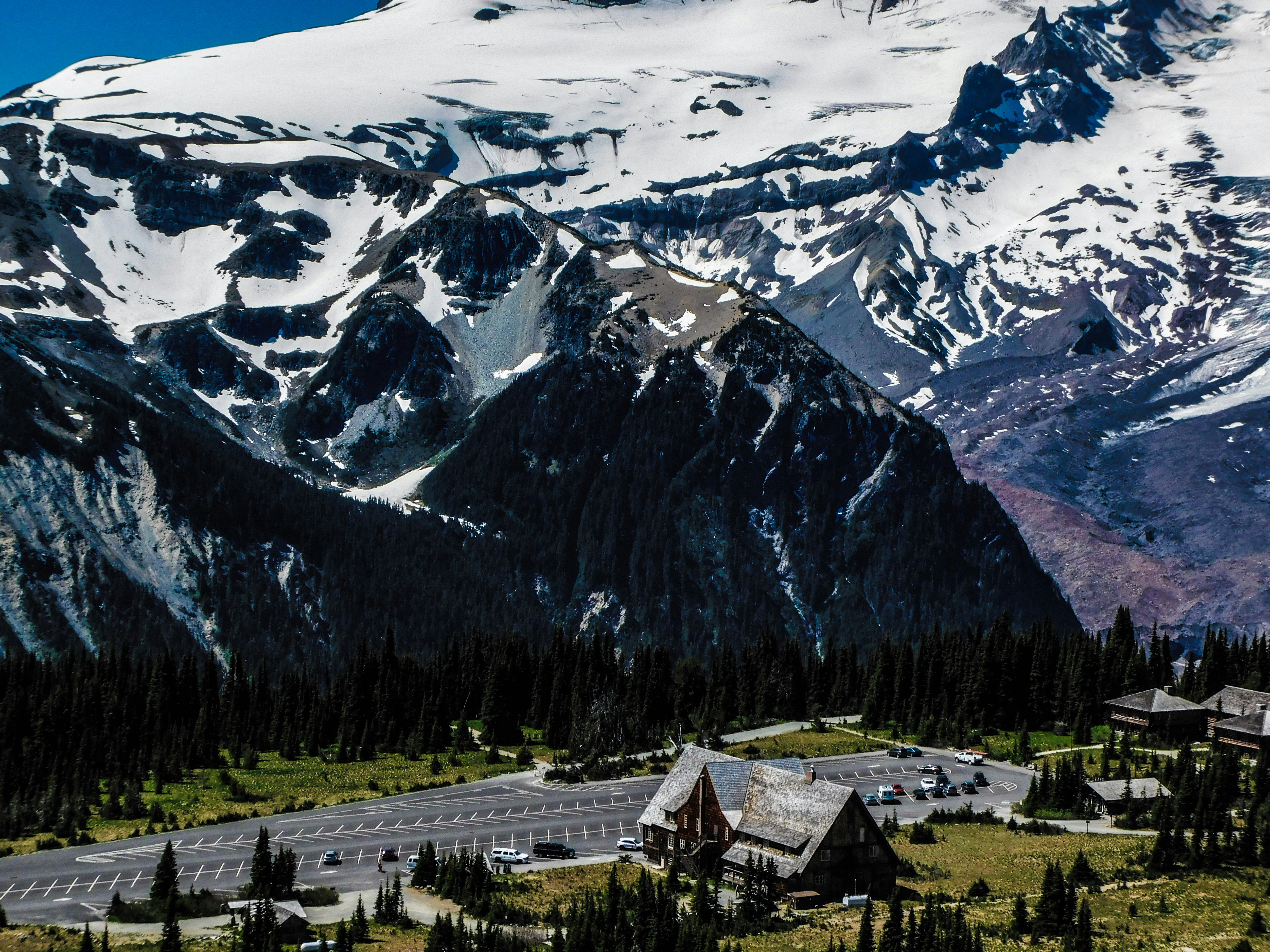 Valley scene with a small wooden chalet beside a winding road, framed by evergreen trees and snow-dusted mountains—a landscape photograph.
