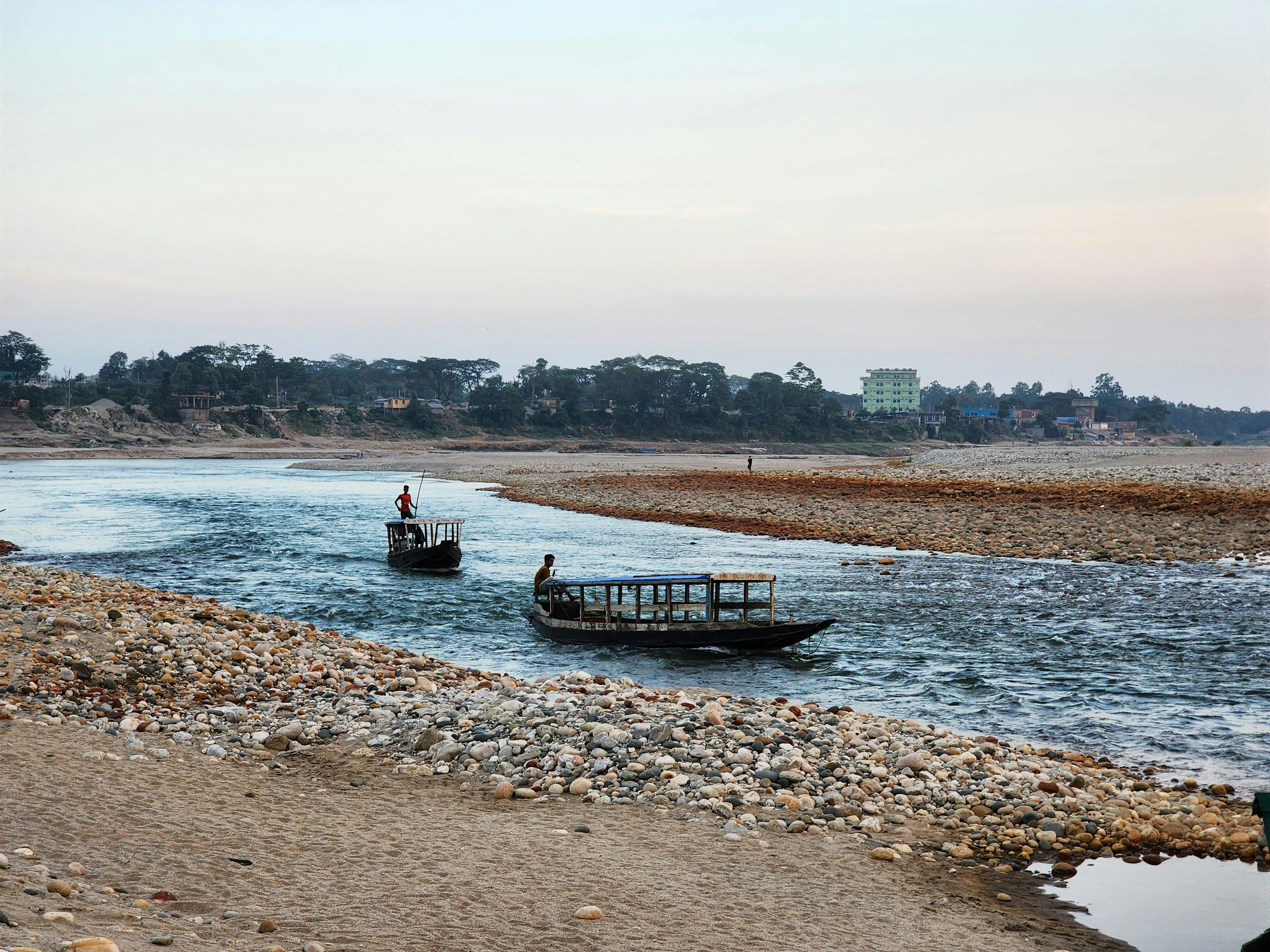Two boats glide along a winding river under a pastel sky, bordered by a rocky shoreline and distant trees.