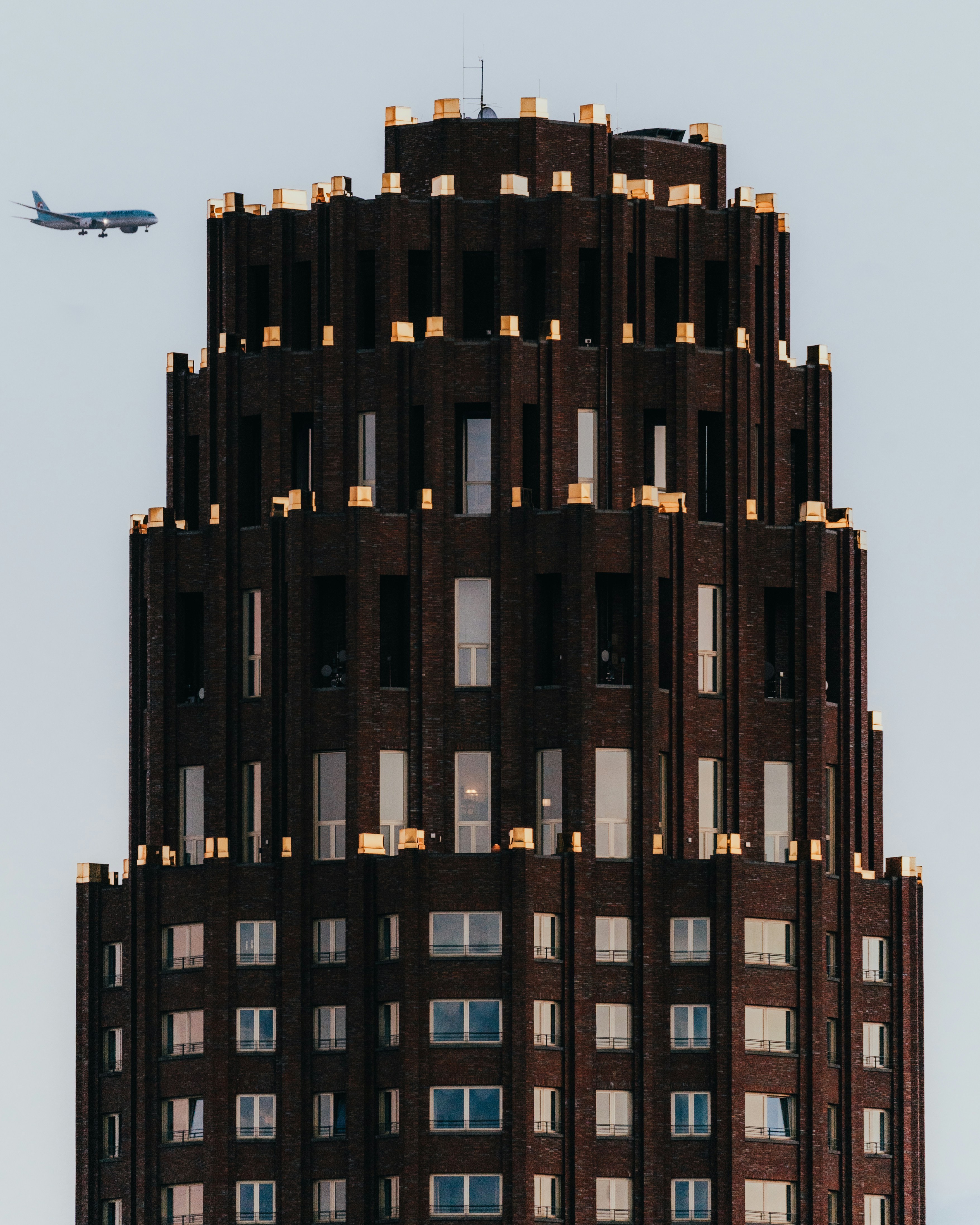 A tall brick Art Deco tower with stepped setbacks rises against a pale blue sky as a small airplane passes to the left, providing a sense of scale.