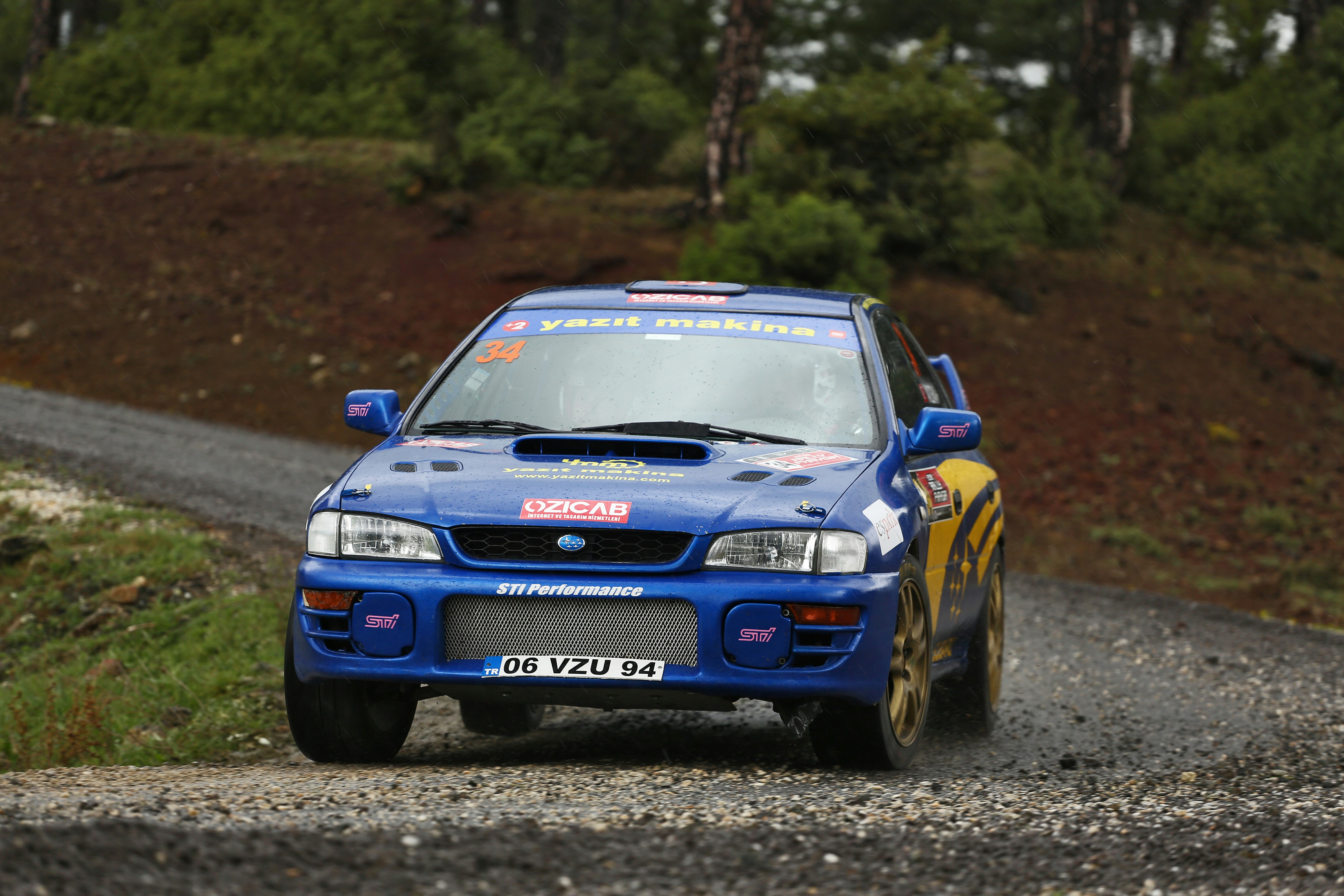 Blue rally car speeding along a winding dirt road in a wooded area.