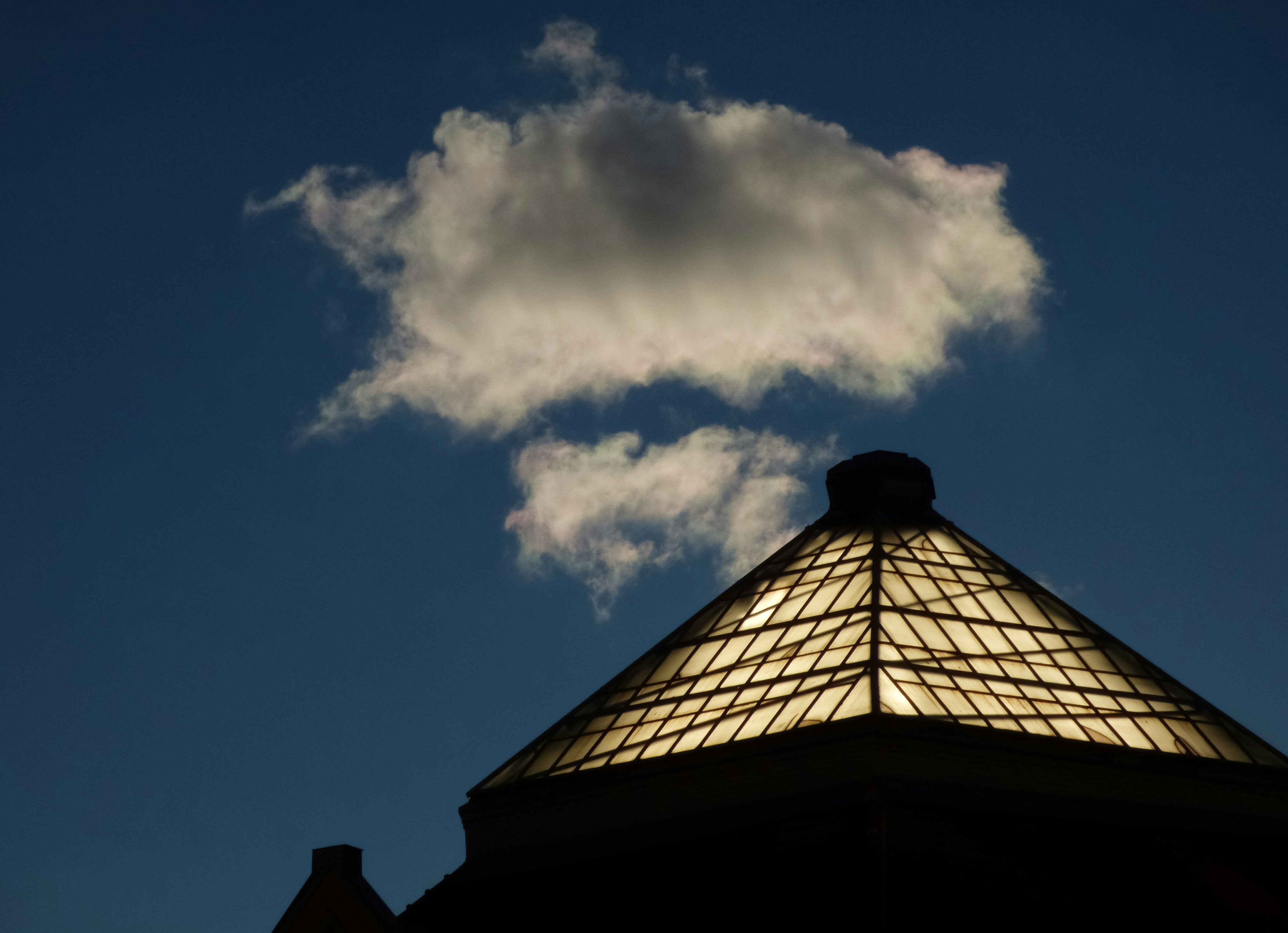 Warm, lit glass pyramid rooftop rises against a deep blue sky with a single cloud.