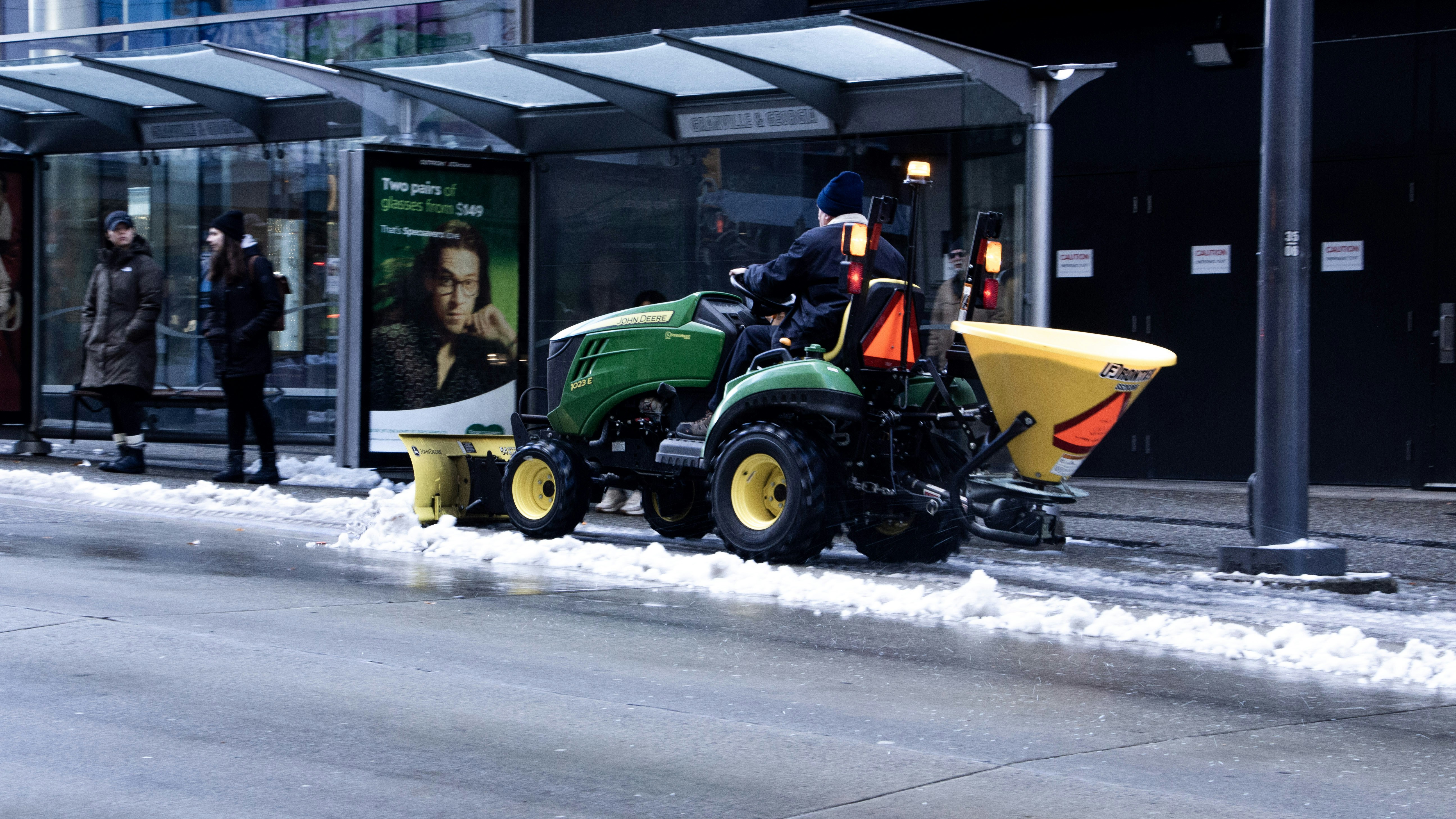 Green Snow-Clearing Tractor Braving a City Street by a Glass-Front Bus Shelter