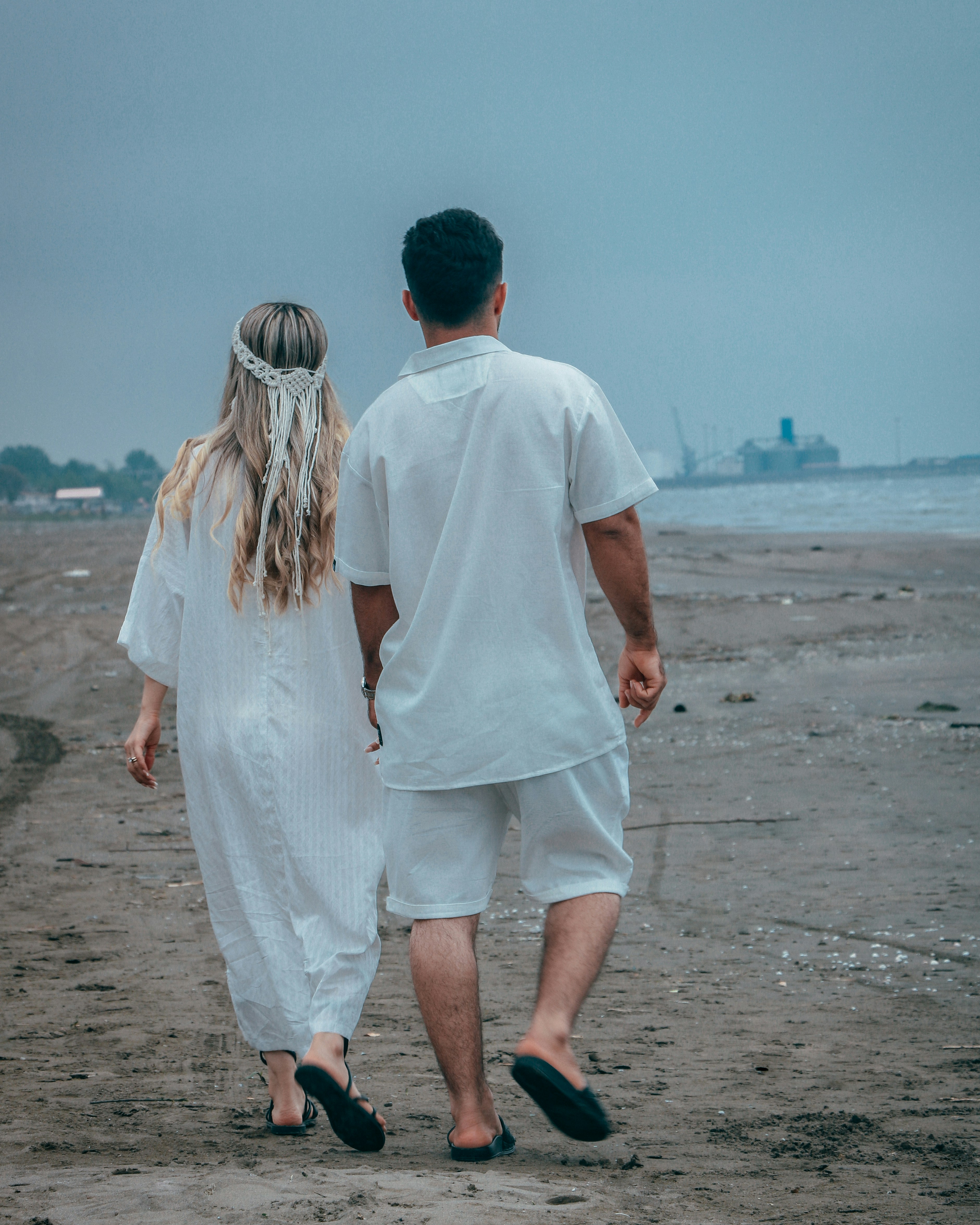 Two people in white clothing walk away along a desolate beach toward a distant shoreline and calm harbor; the scene emphasizes mood over detail, with soft light and a subdued blue-gray horizon. The image captures a candid, contemplative moment.