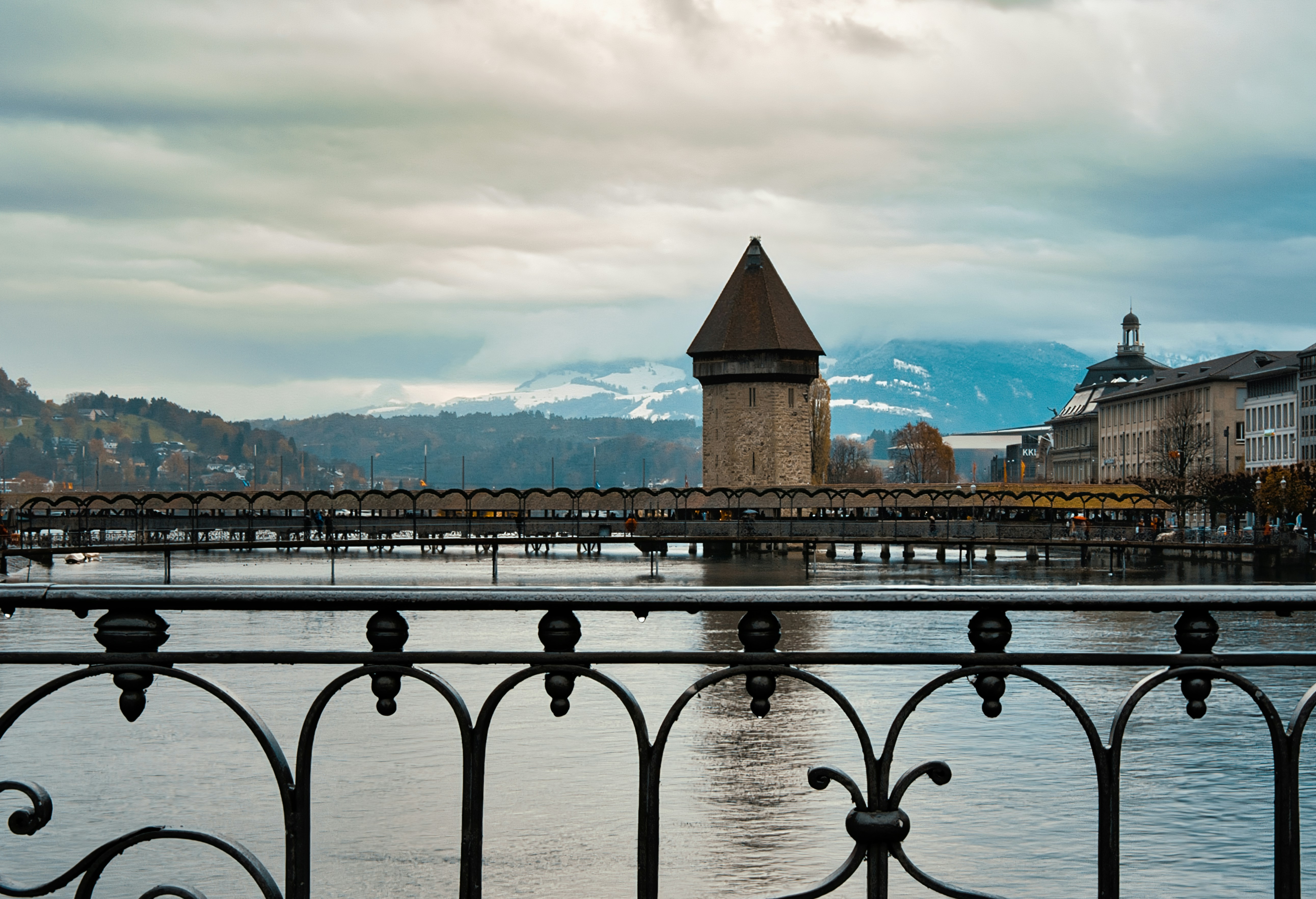 Historic water tower standing by the serene lake, framed by an ornate wrought-iron railing. The backdrop features distant mountains under a cloudy sky.