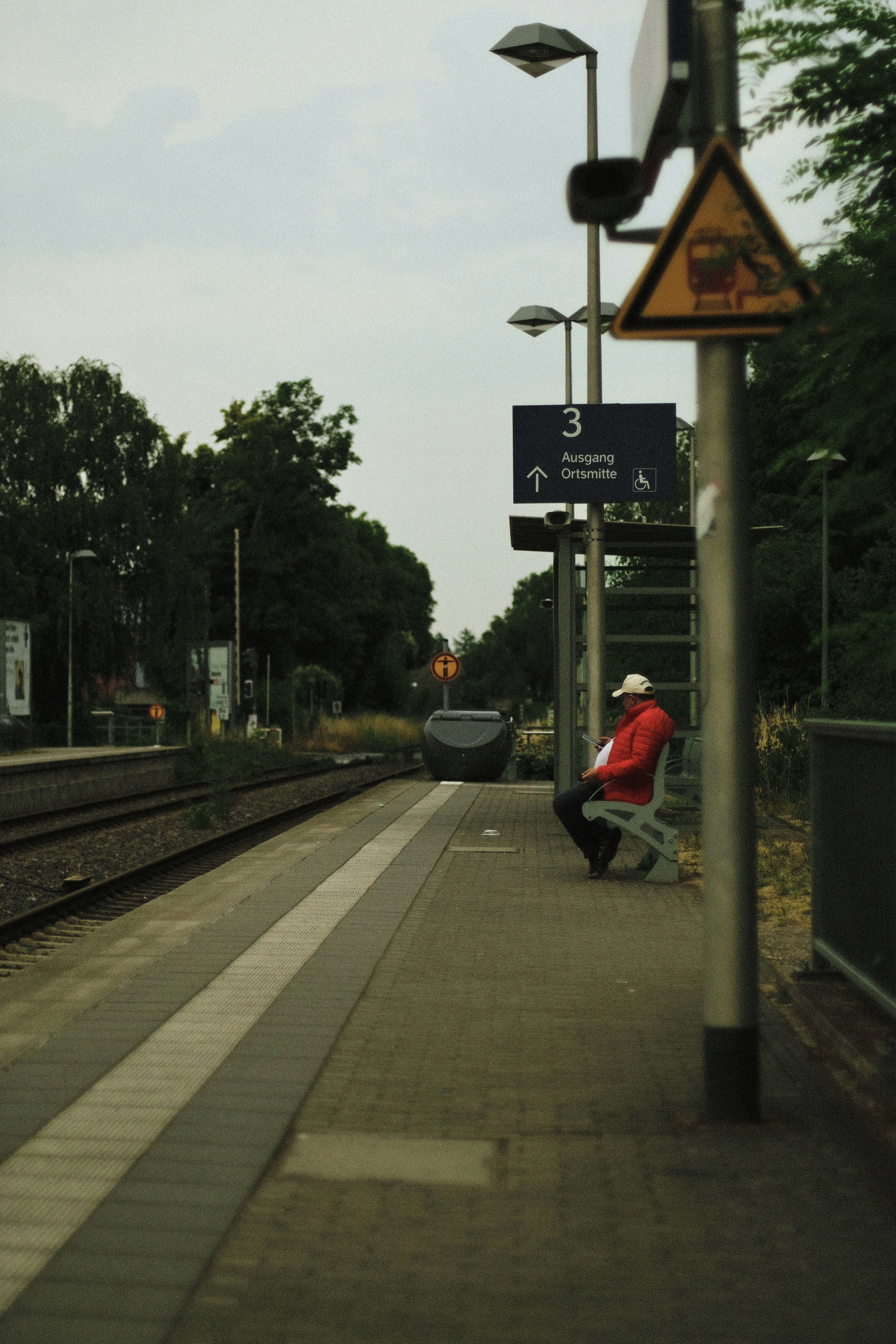 A solitary figure in a red jacket sits on a bench at a train station, awaiting the arrival of a train. The scene is framed by lush greenery and urban infrastructure.