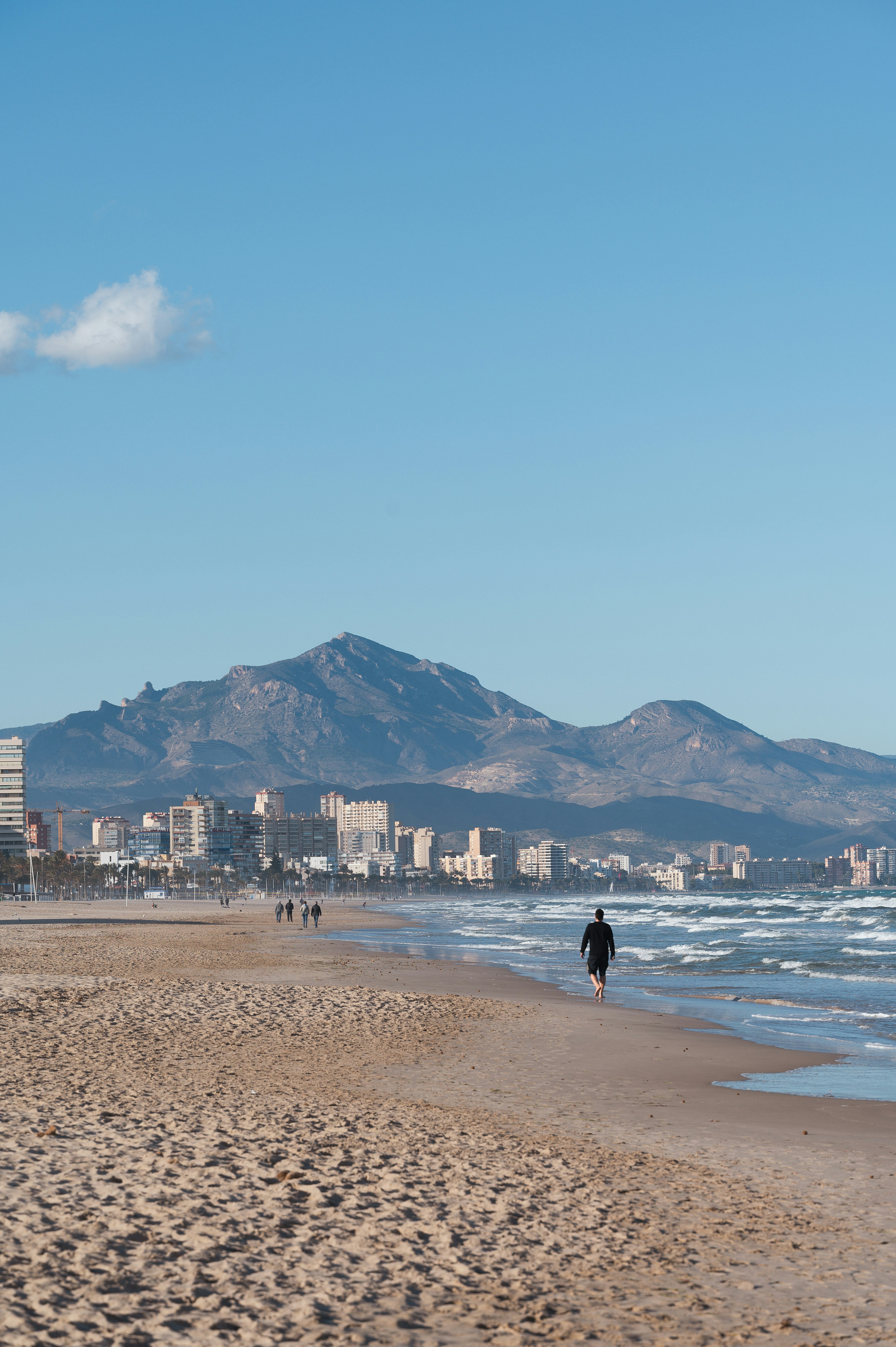 A solitary figure walks along a sandy beach with waves lapping at their feet, framed by a backdrop of distant mountains and cityscape.