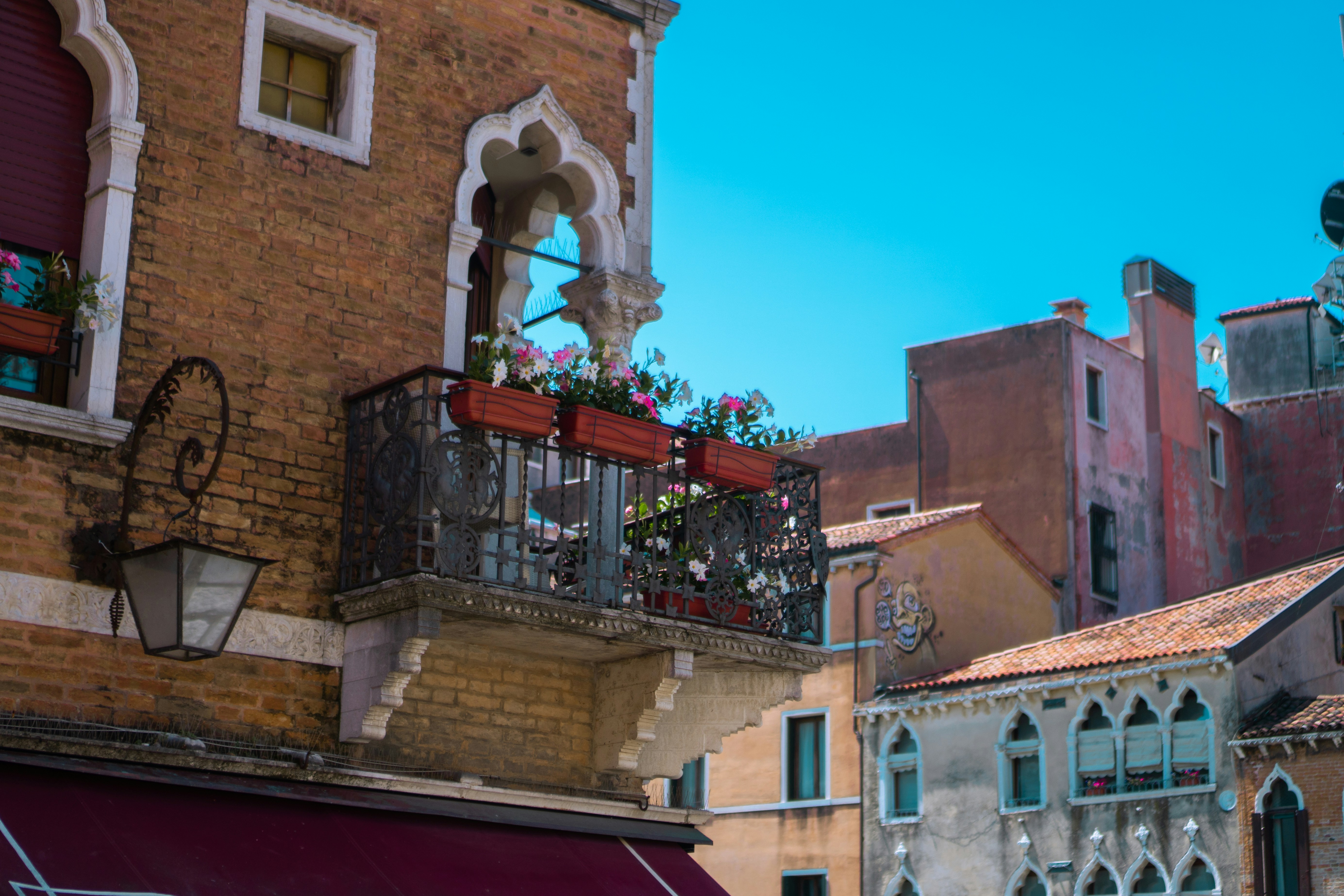 This image captures a picturesque Venetian balcony adorned with vibrant flower boxes, set against a backdrop of warm, textured brick and pastel-hued buildings. The daylight casts a soft, natural glow, enhancing the vivid colors of the flowers and the rich tones of the architecture. The intricate ironwork of the balcony adds a touch of elegance, making the scene both inviting and visually striking.
