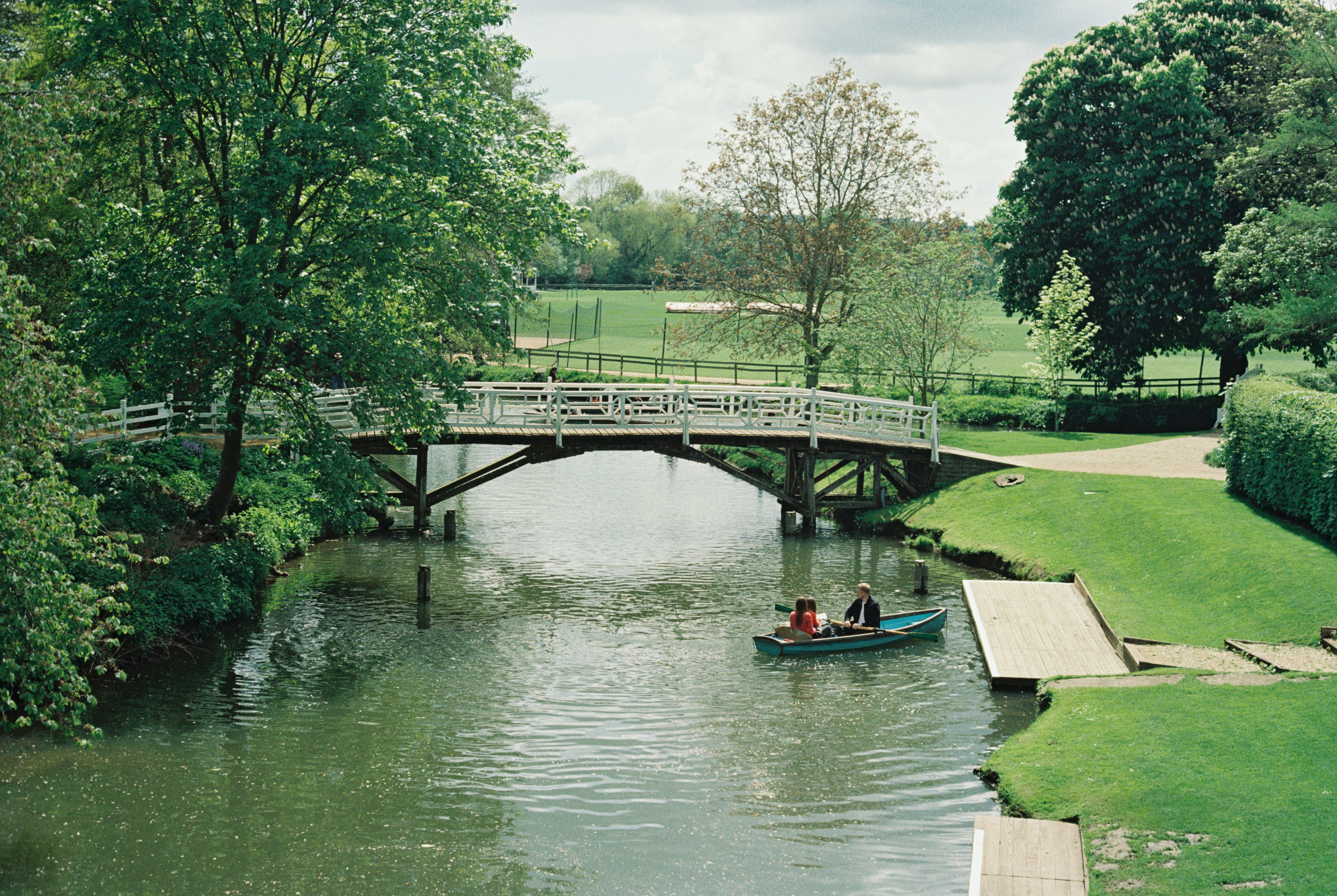 A tranquil park photograph of a small rowboat on a calm river passing beneath a wooden arched bridge, surrounded by lush trees and manicured lawns.