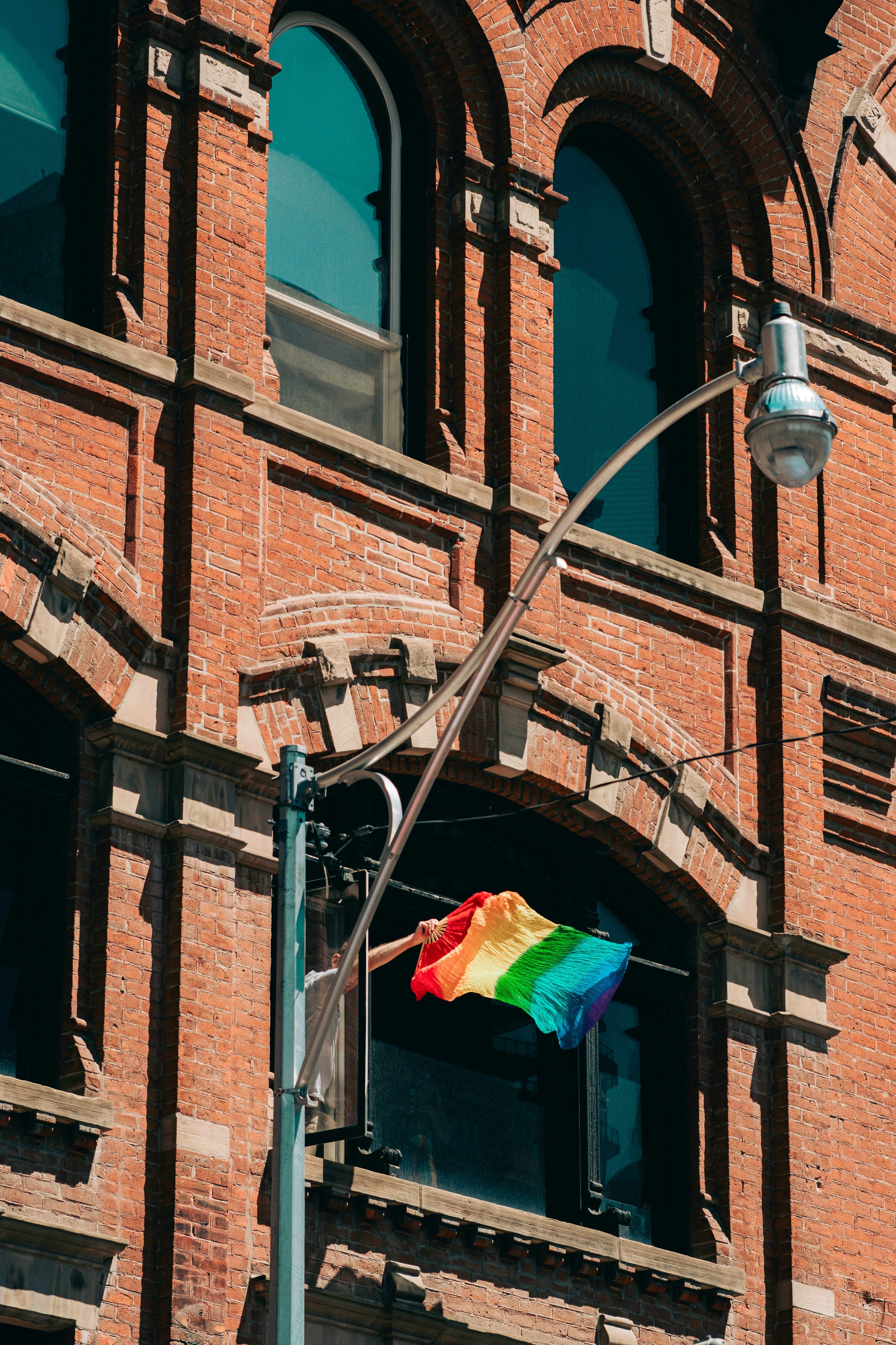 Rainbow Flag Drapes from a Curved Streetlamp Before a Red Brick Facade