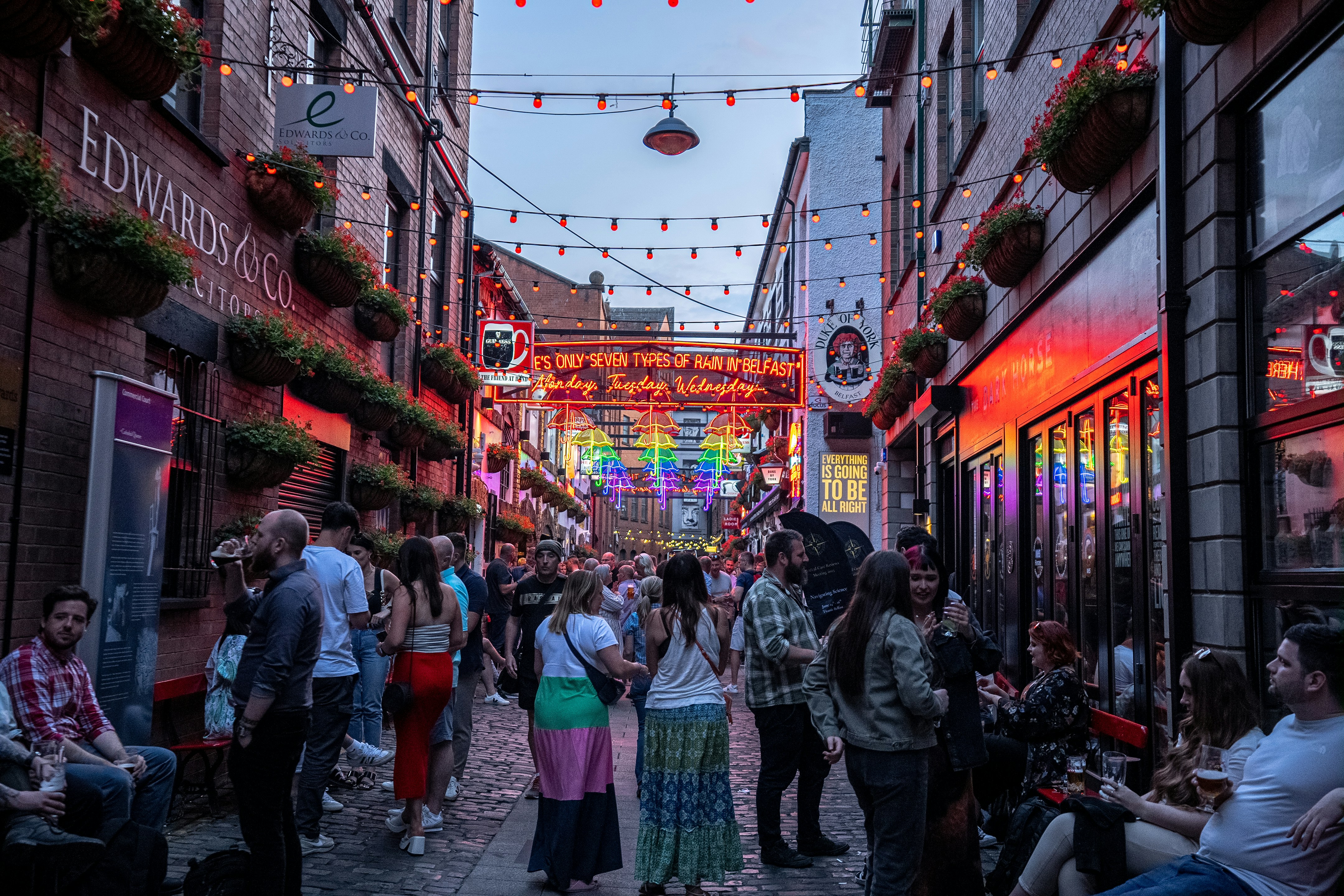 Bustling alleyway filled with people enjoying the lively atmosphere, adorned with colorful neon lights and flower baskets. The scene captures the essence of urban nightlife.