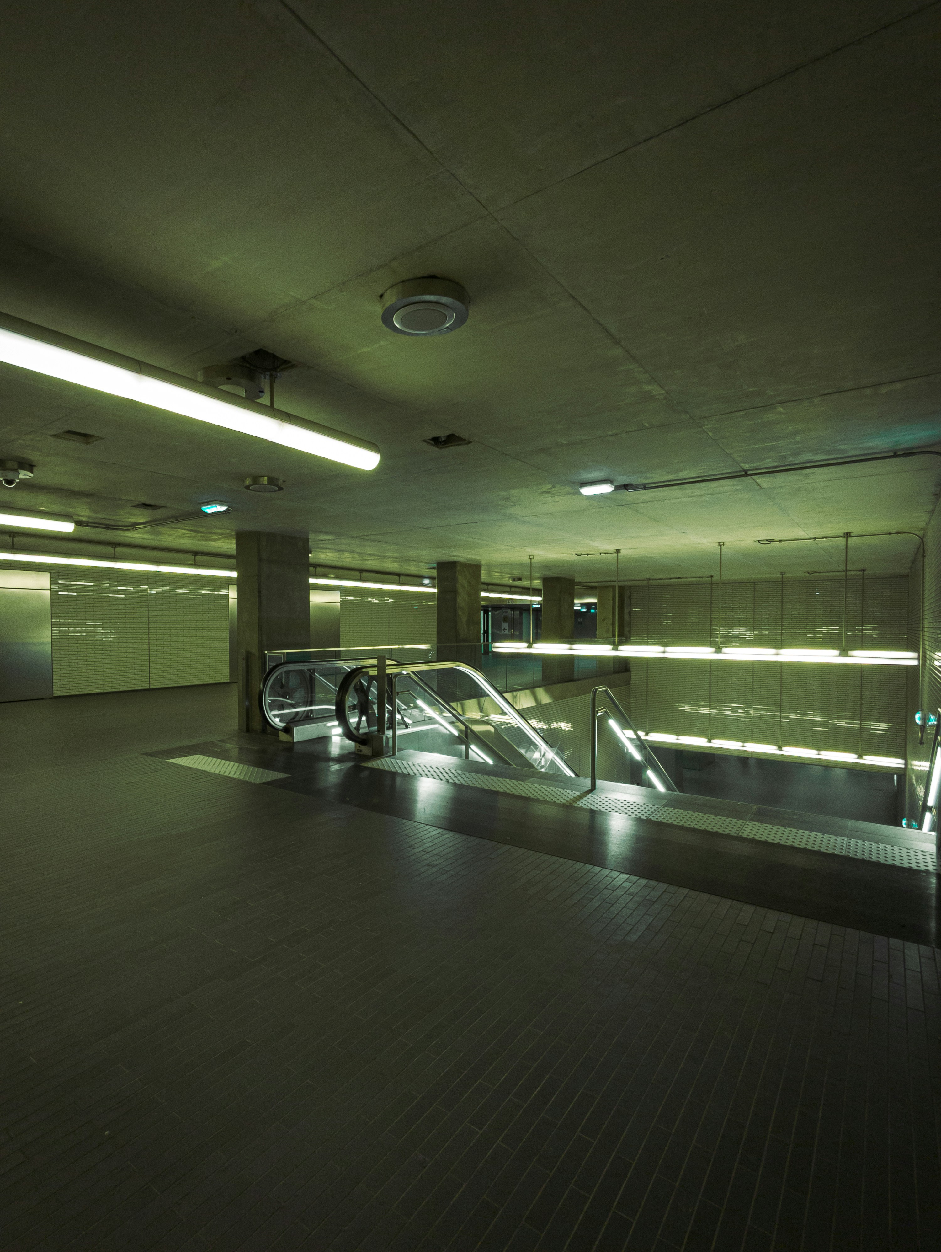 An empty underground station hall with a bright escalator and glass walls, bathed in cool artificial light. The scene emphasizes quiet architectural minimalism rather than human activity.