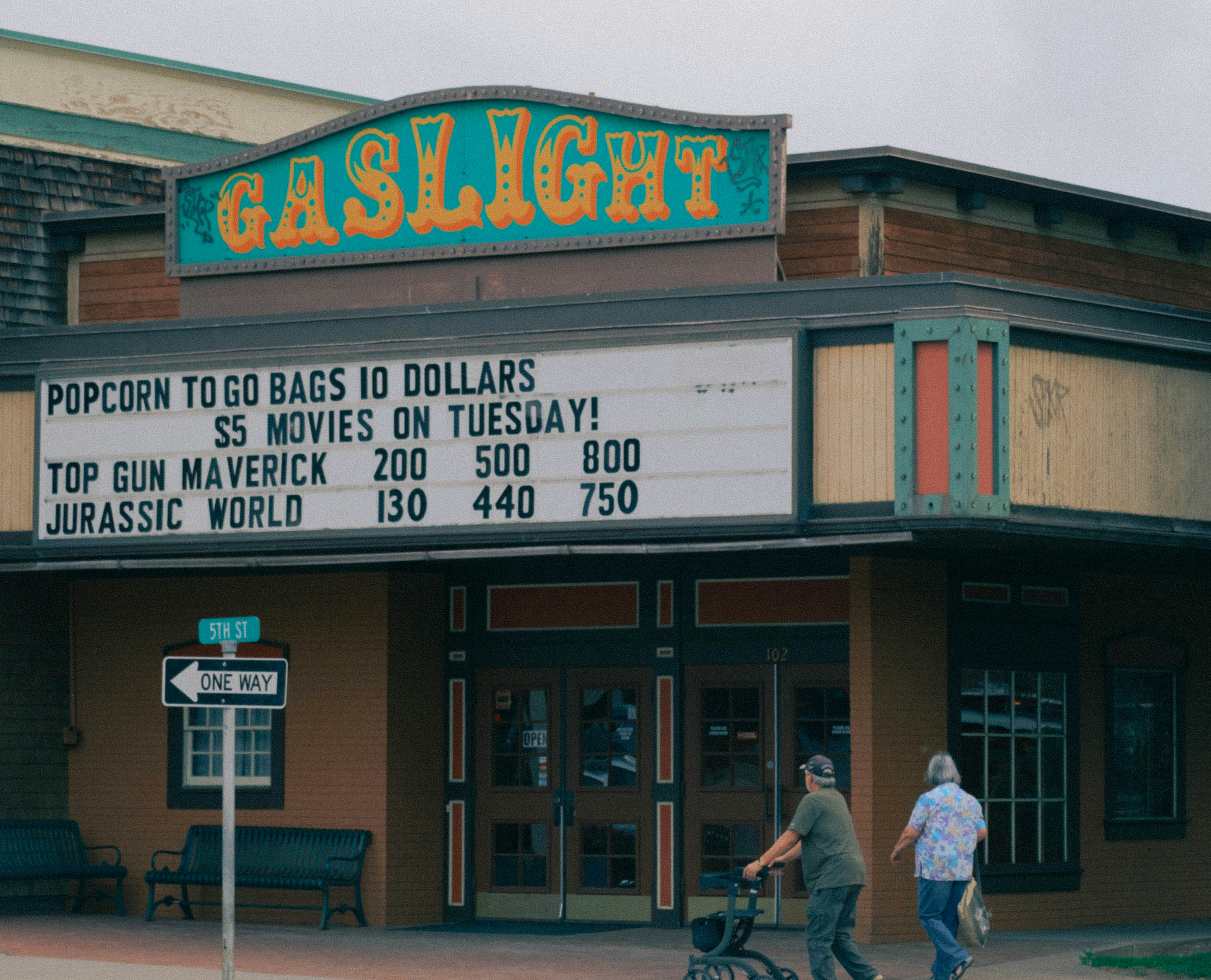 Vintage theater with marquee displaying movie showtimes, featuring pedestrians passing by on a cloudy day.