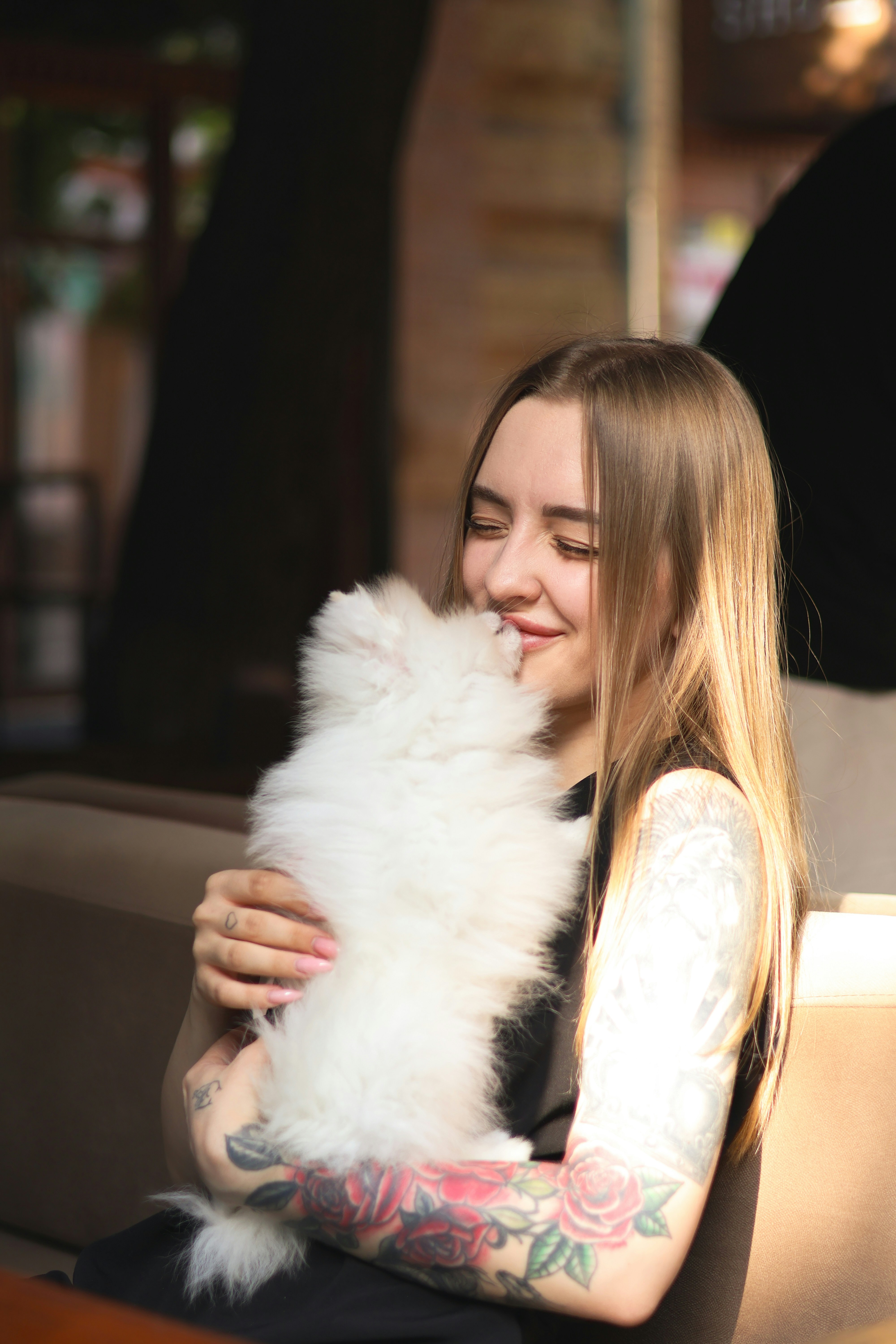 Photograph of a tattooed woman in a cafe cuddling a fluffy white companion, bathed in warm light.