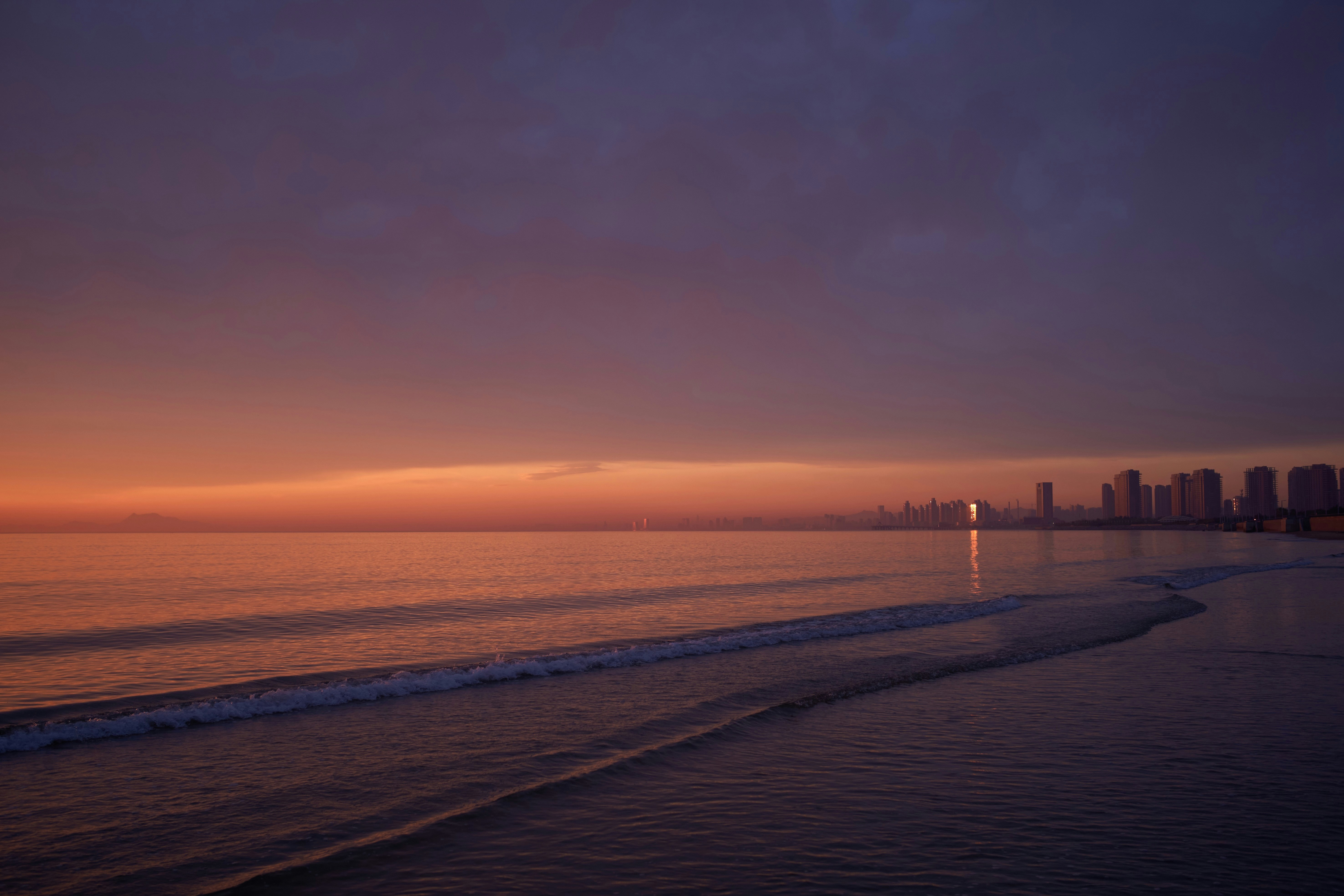 A sunset view of a beach with a city in the distance photo – Free Sky ...