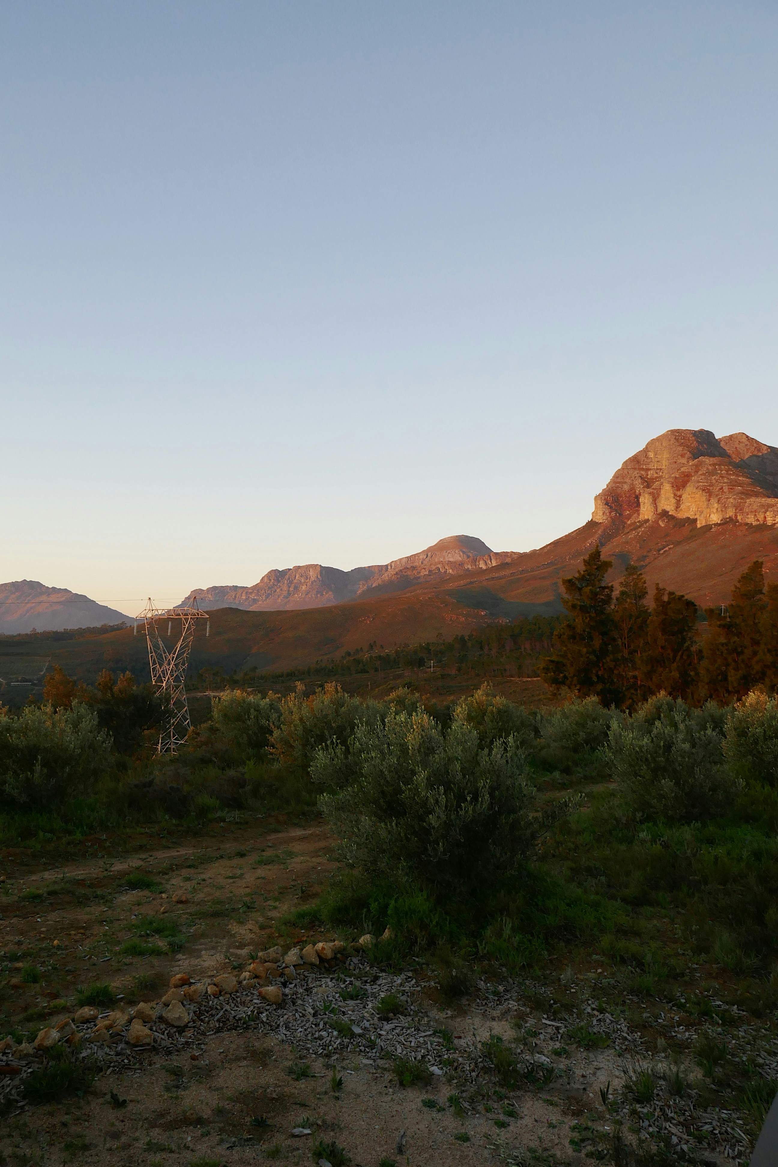 Desert landscape at golden hour with a sunlit mountain ridge on the right, sparse shrubs in the foreground, and a transmission tower toward the left.
