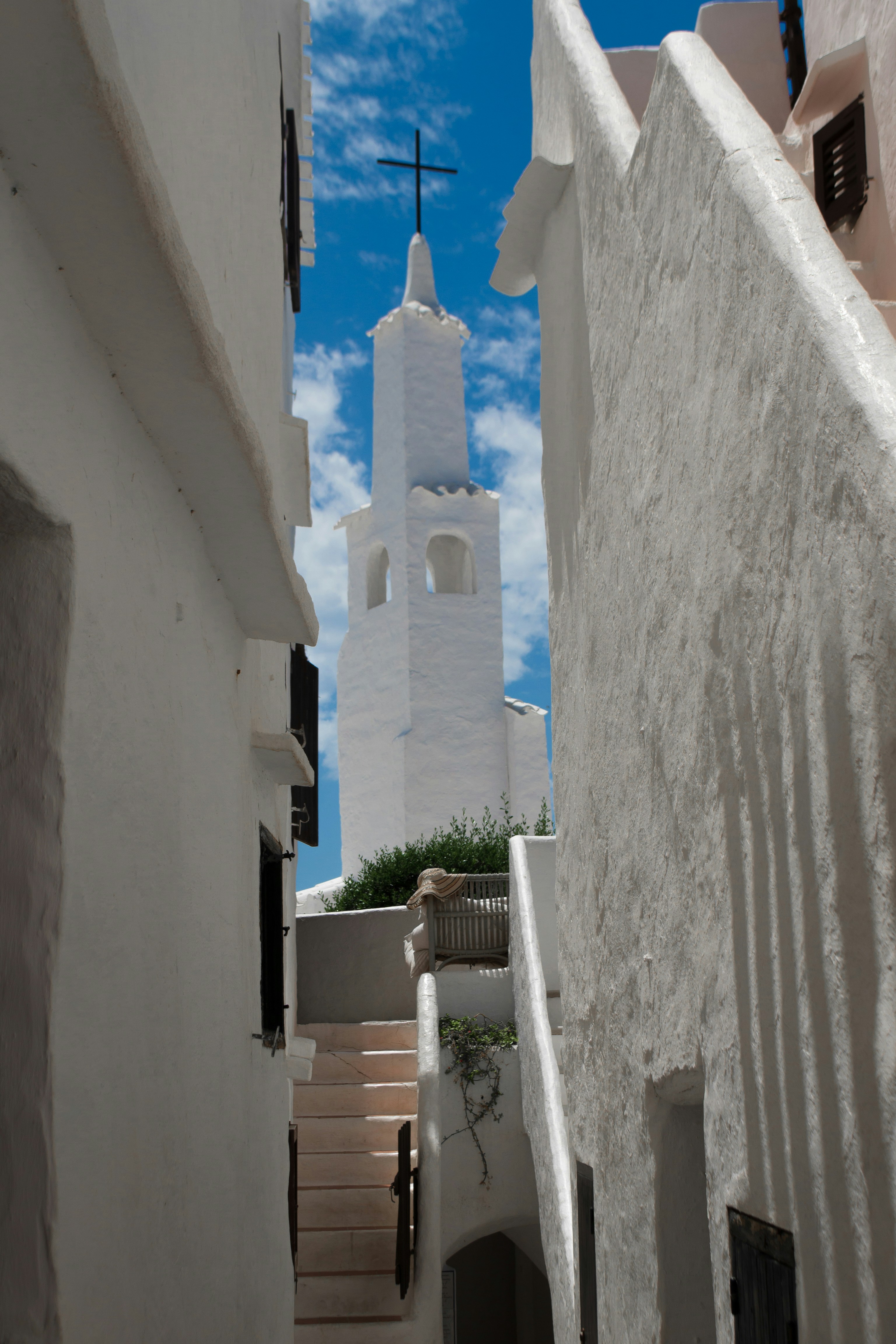 A serene alleyway framed by whitewashed walls leads to a church tower under a bright blue sky, showcasing the charm of Mediterranean architecture.