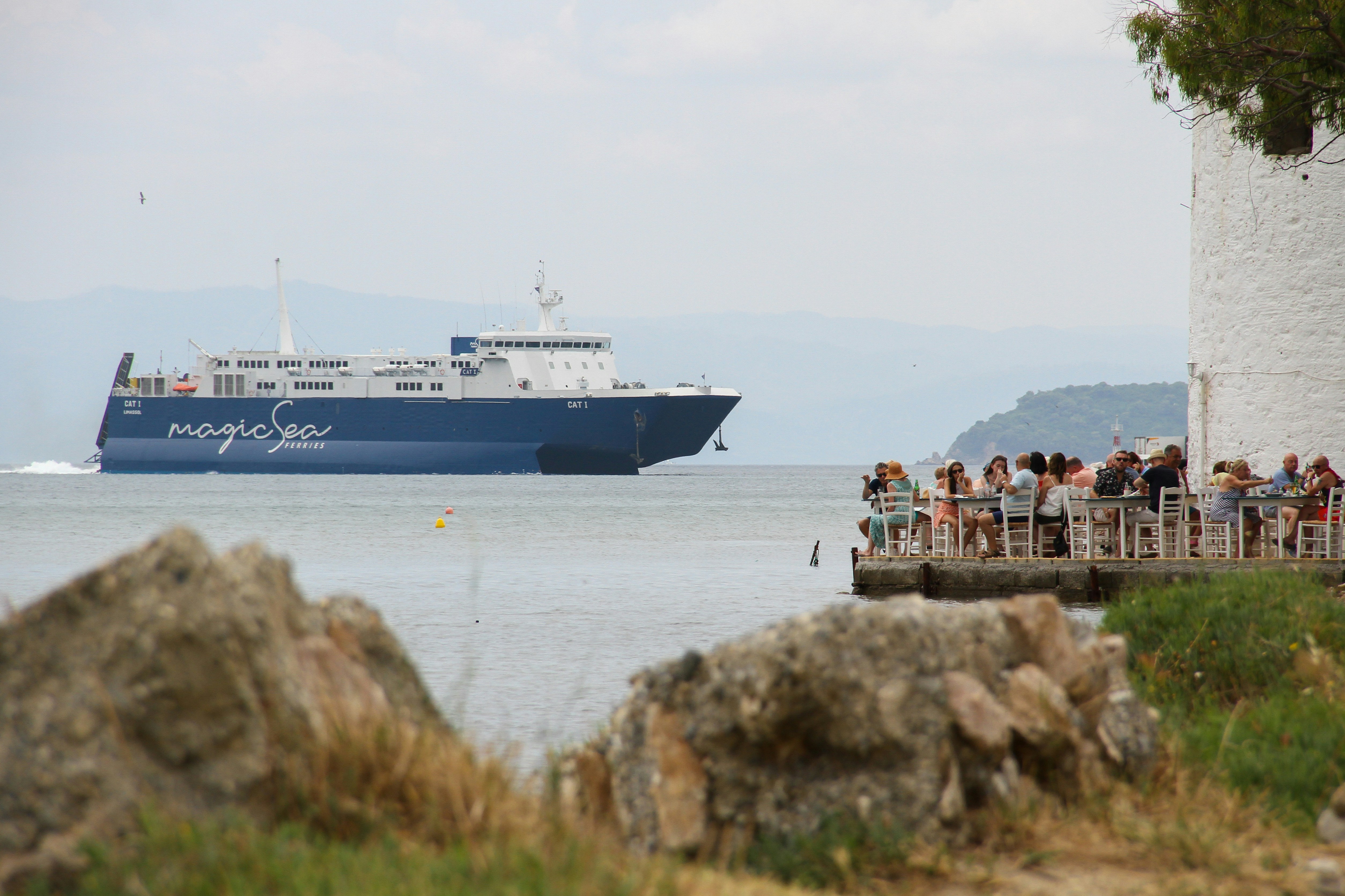 Blue ferry gliding past a sunlit cliffside café