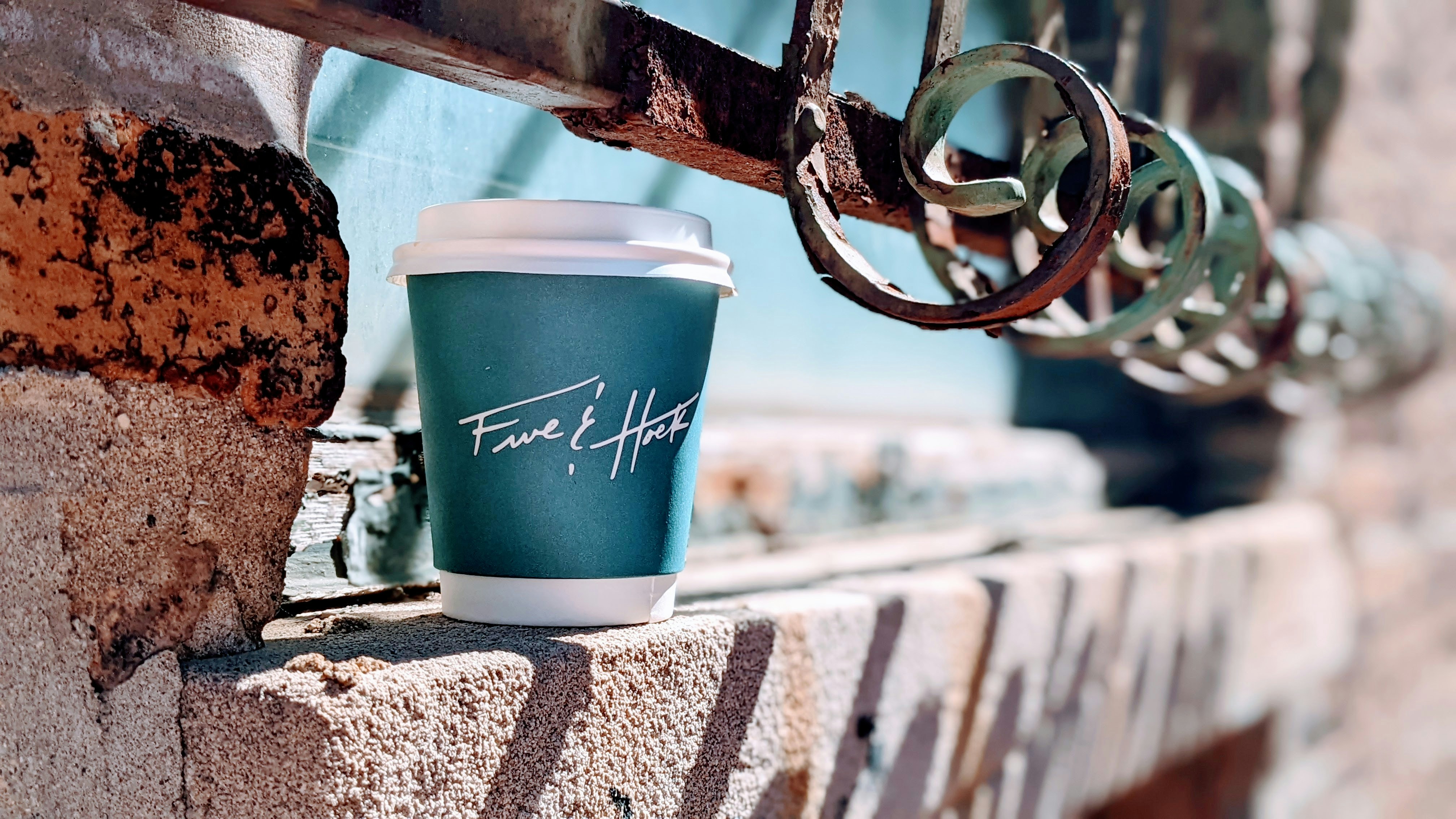 Teal coffee cup with a handwritten logo rests on a rustic stone ledge, framed by an ornate metal gate under bright sunlight.
