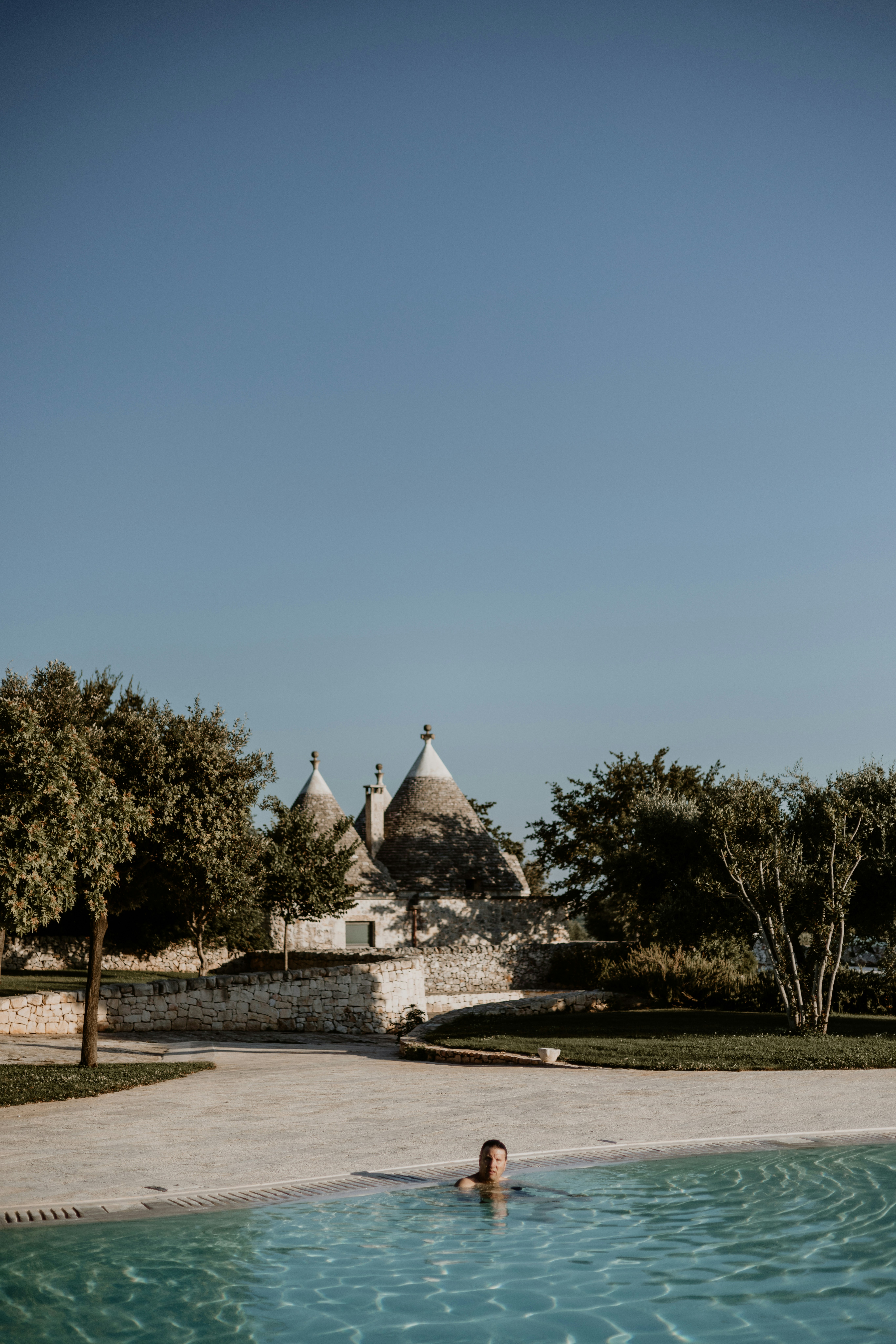 Person swimming in a pool with historic trulli buildings in the background and surrounded by trees.