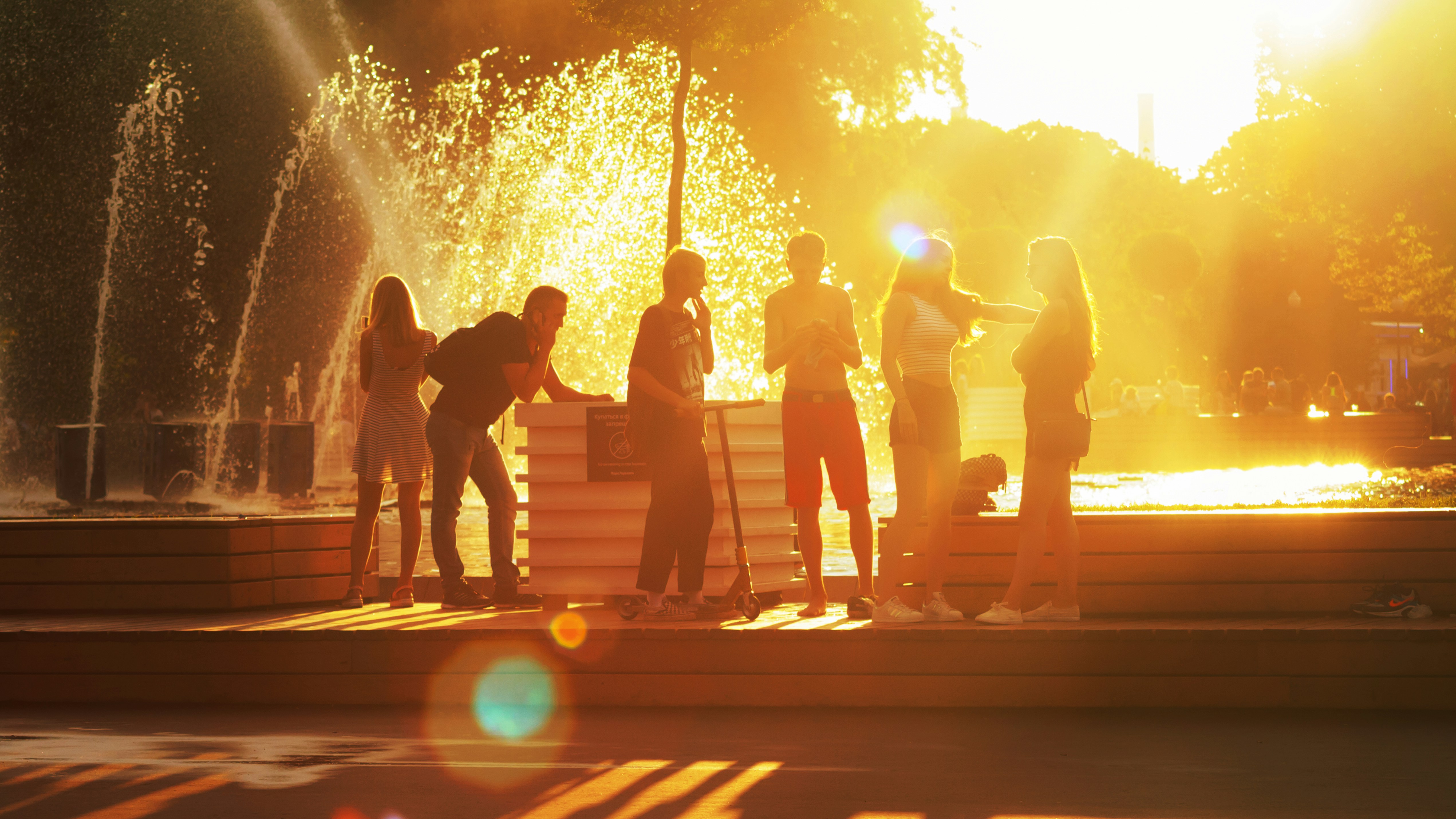 Late-afternoon silhouettes gather around a sunlit fountain, warm backlight turning figures into dark shapes while water glows. A public plaza scene highlighting group interaction and architectural steps.
