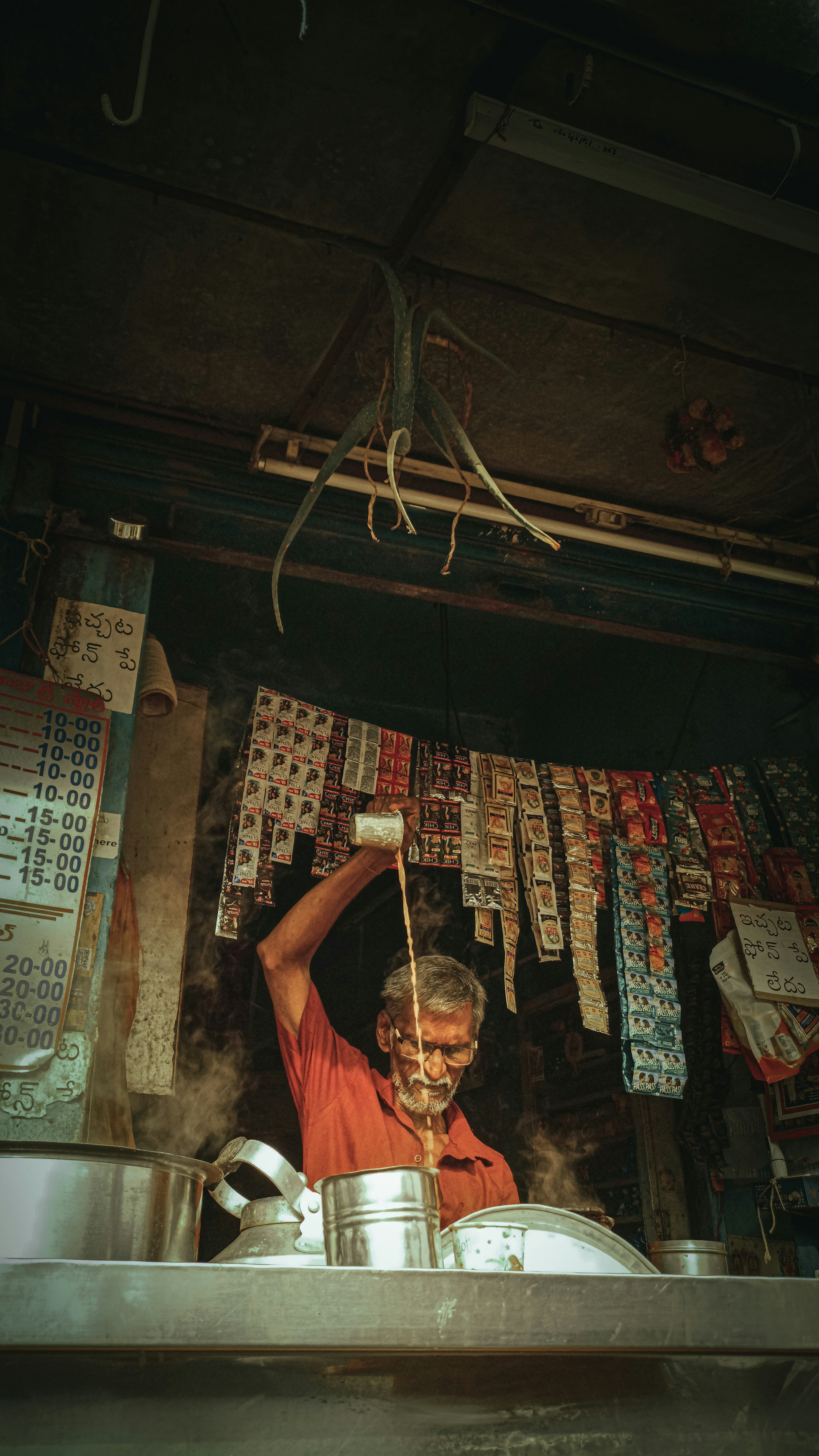 Street vendor pours liquid from a ladle into a pot as steam rises, with colorful market cards and signs in the background. The scene captures everyday hustle in a compact, busy market kitchen.
