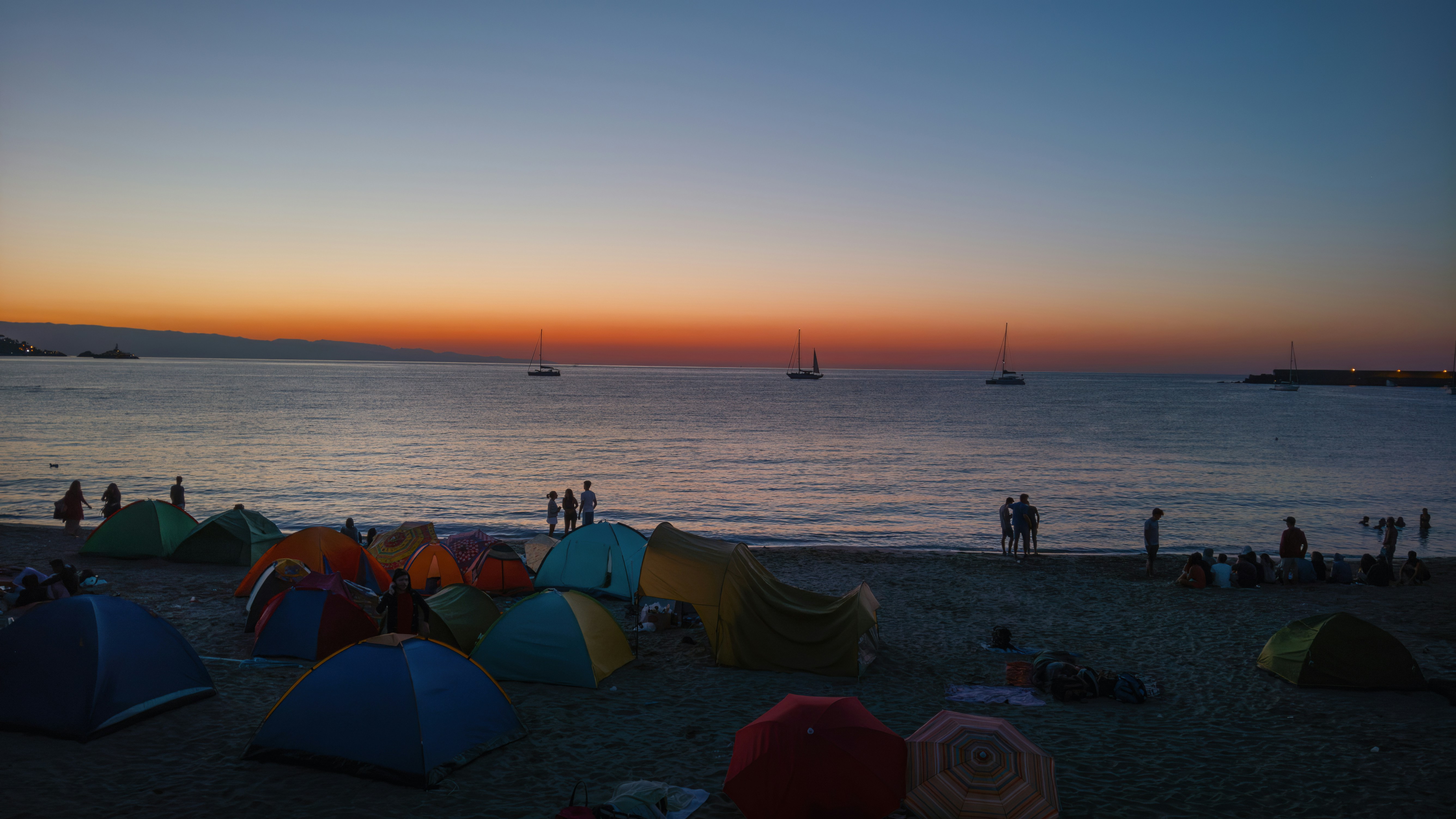 Colorful tents line a crowded beach as silhouettes gather along the shoreline while sailboats drift on a warm sunset horizon.