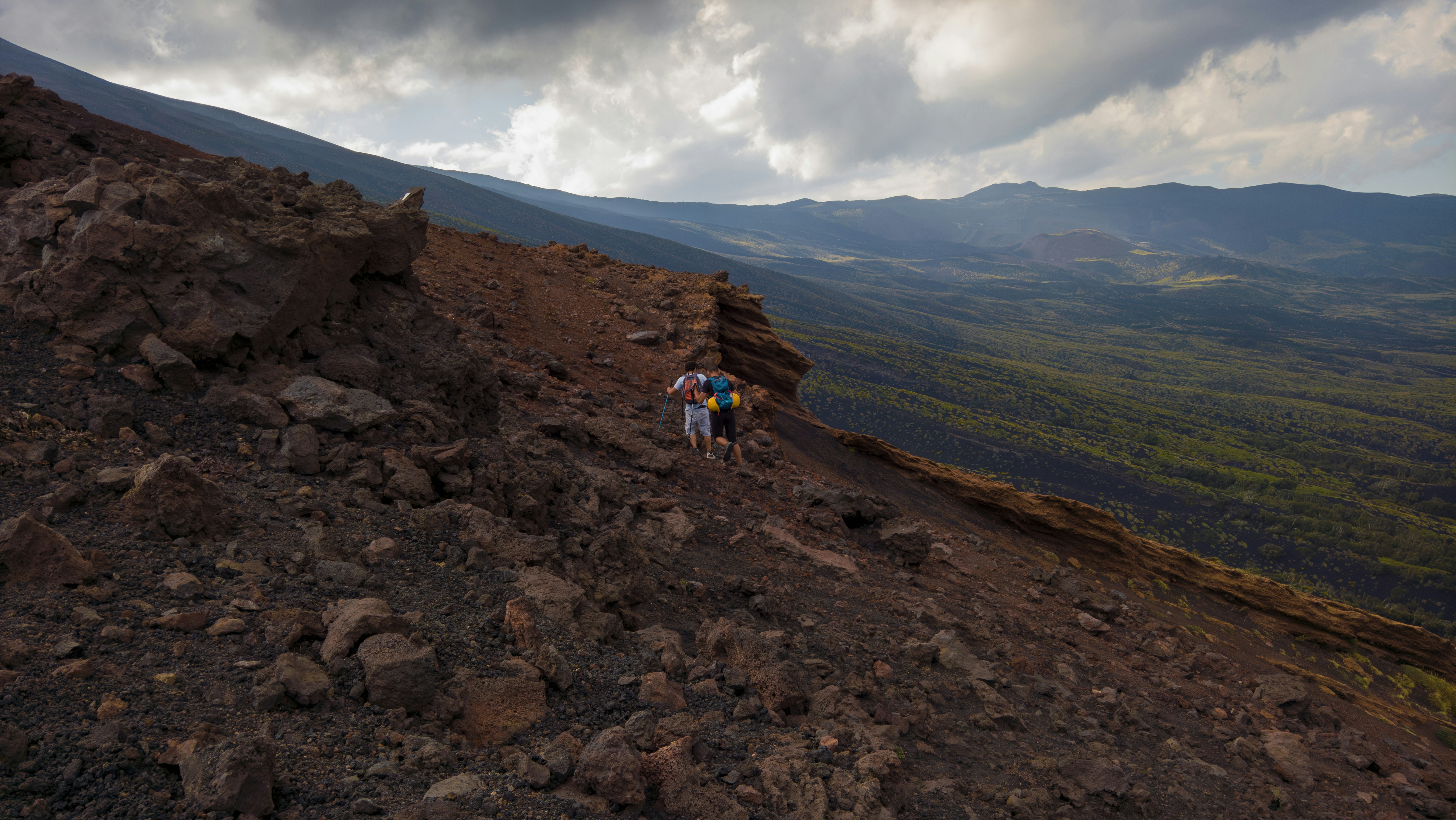 Hikers traverse a rugged volcanic landscape beneath dramatic clouds.