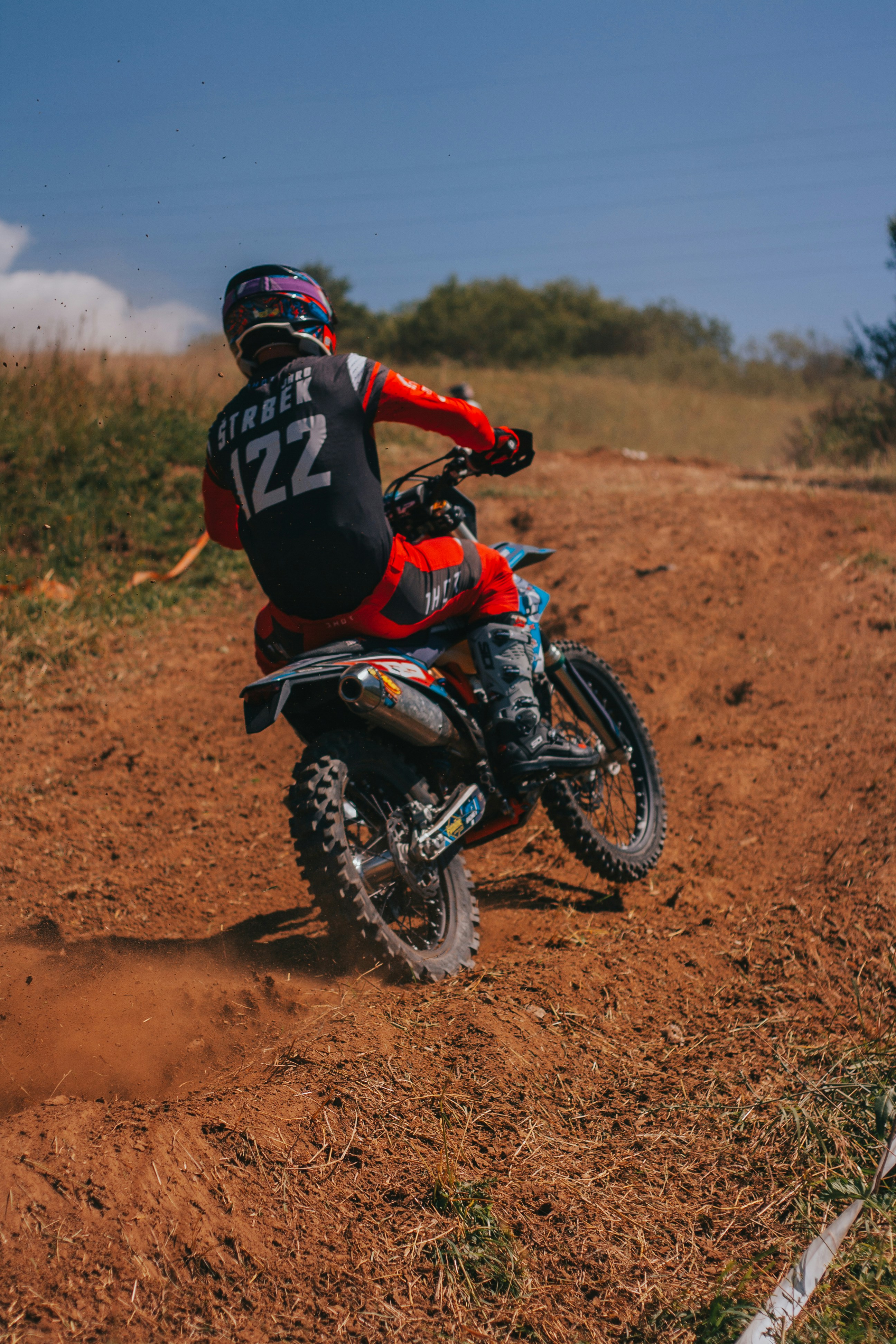 Motorcyclist in red gear rides up a dirt trail under a clear blue sky.