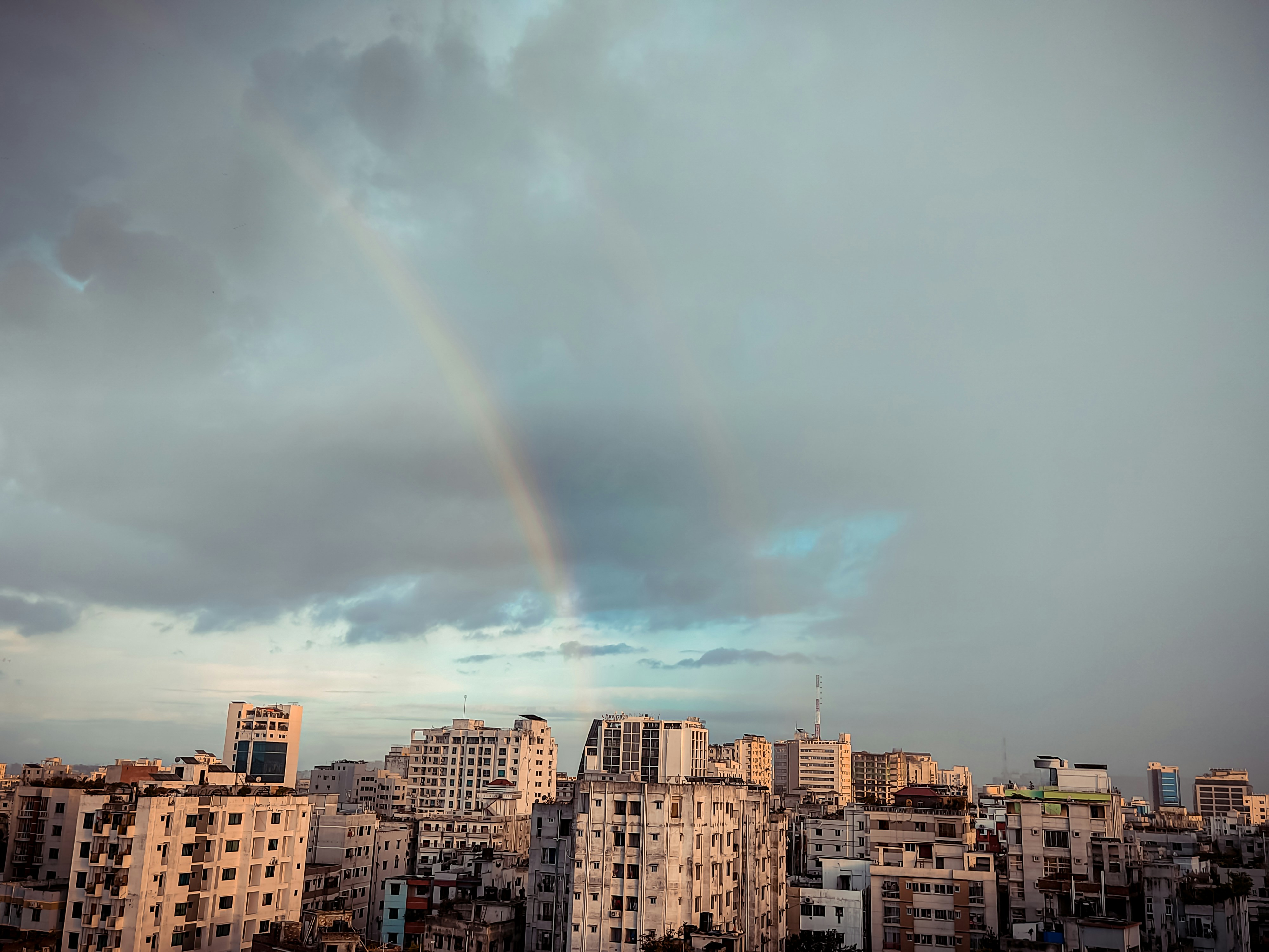 City skyline at dusk with a soft rainbow arching over low-rise apartments, capturing urban calm after rain.