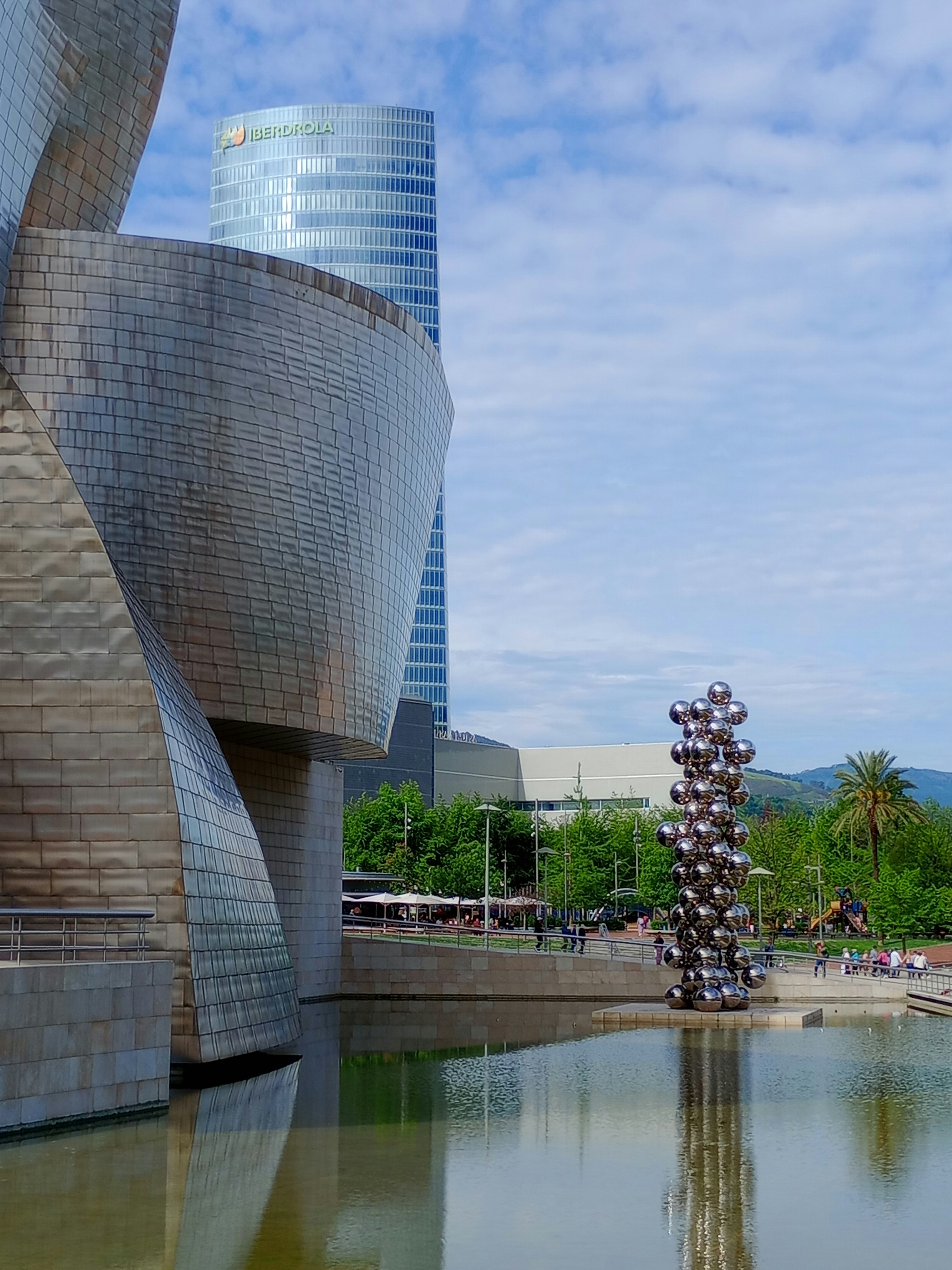 Architectural marvel with metallic curves beside a tranquil waterway, featuring a unique sculpture in the foreground. The scene captures the harmony between nature and urban design.