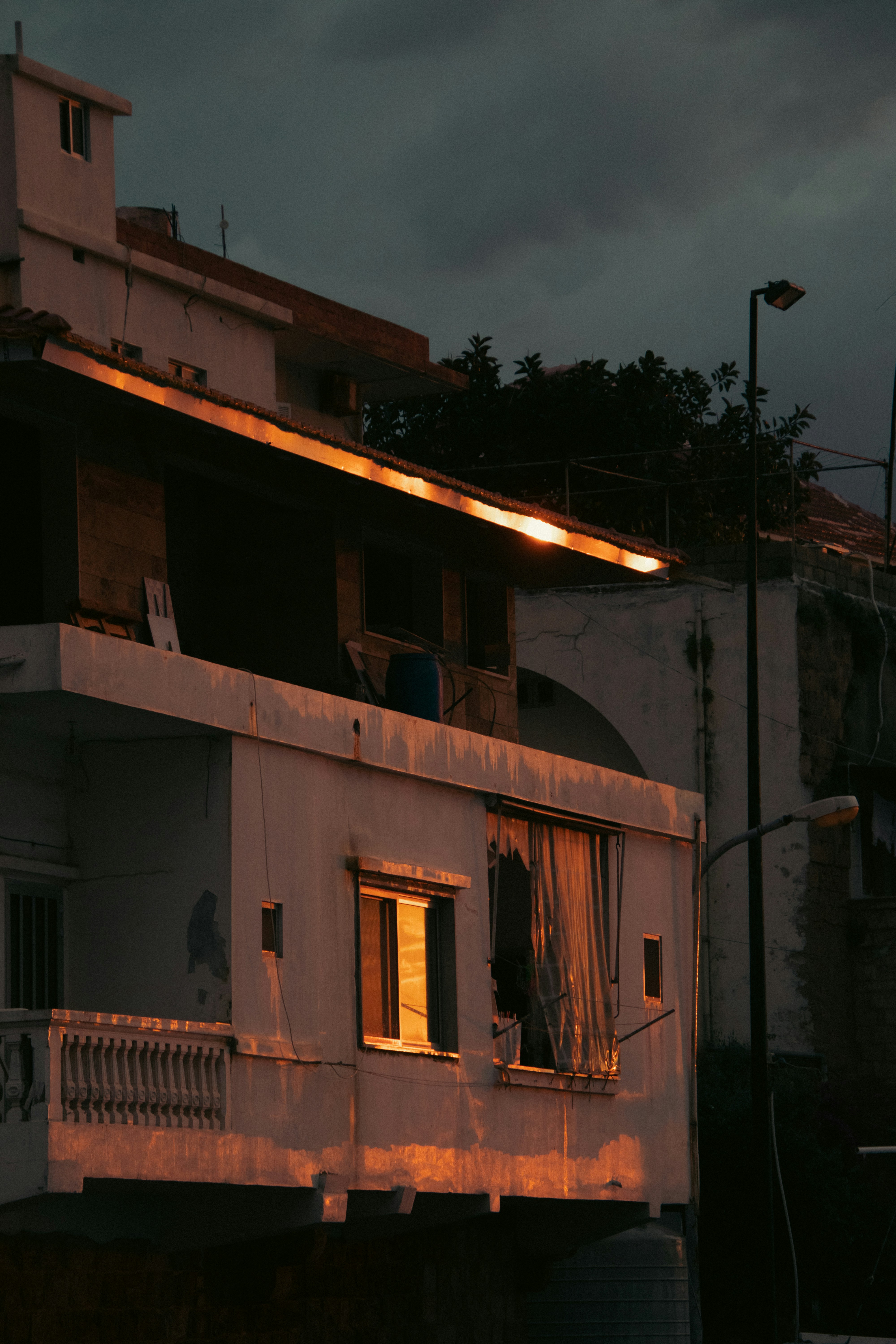 A weathered building reflects warm evening light, highlighting its textures and a window with flowing curtains. The scene conveys a sense of urban solitude.