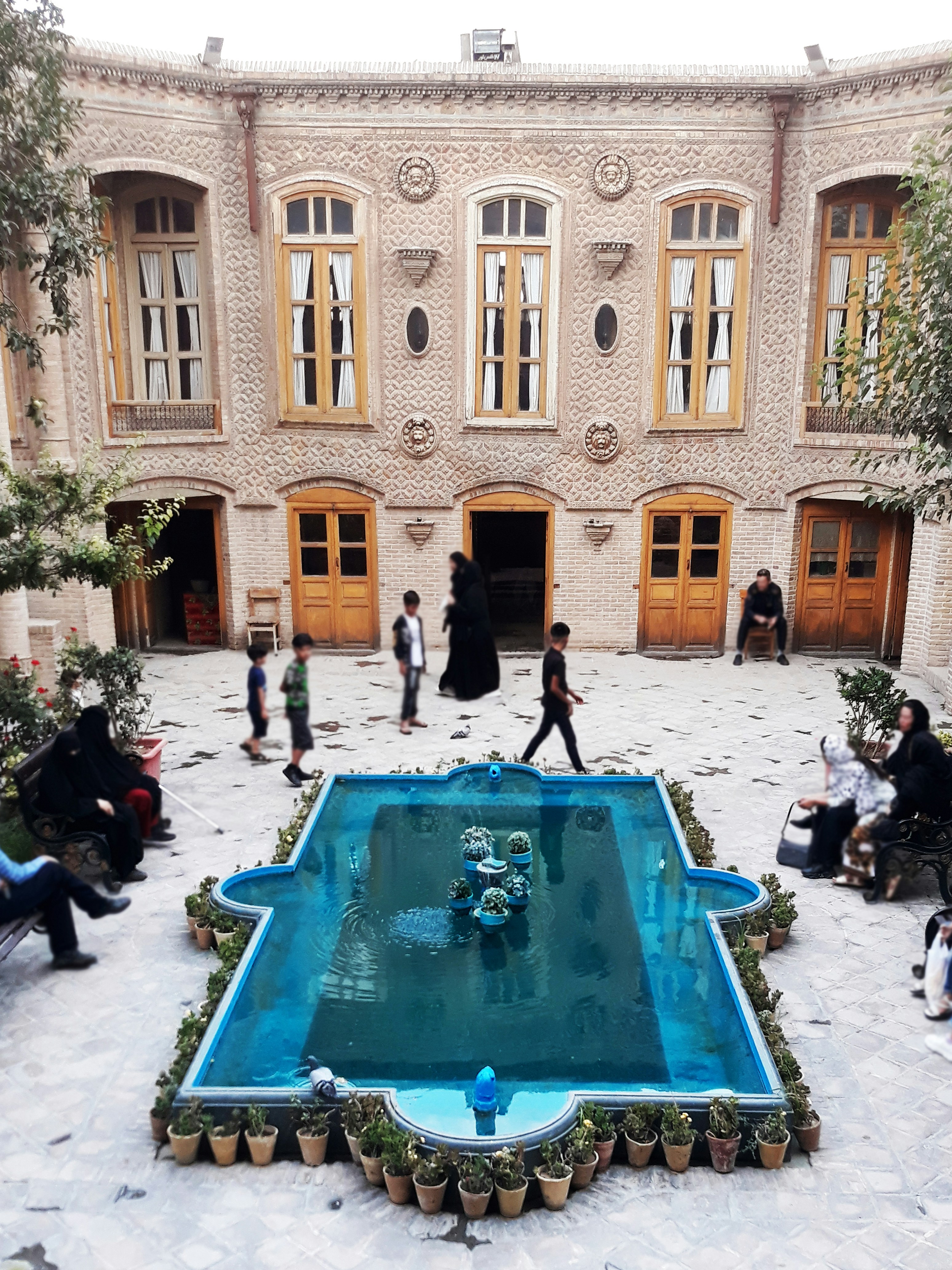 Photograph of a brick courtyard with a central blue fountain surrounded by potted plants. People mingle along the edges, capturing a moment of daily life.