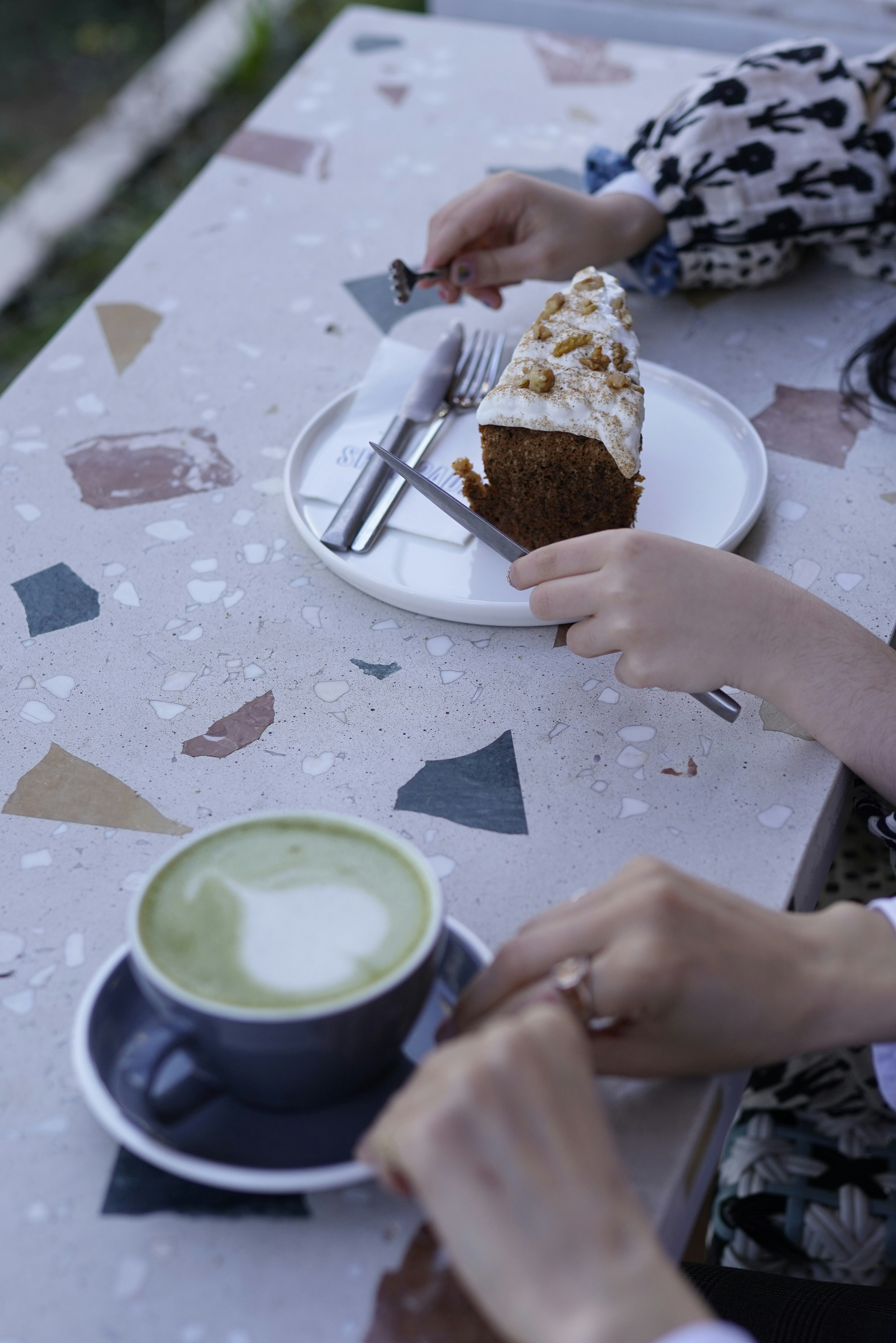Slice of caramel-cream cake on terrazzo table, framed by a poised hand and a green matcha latte