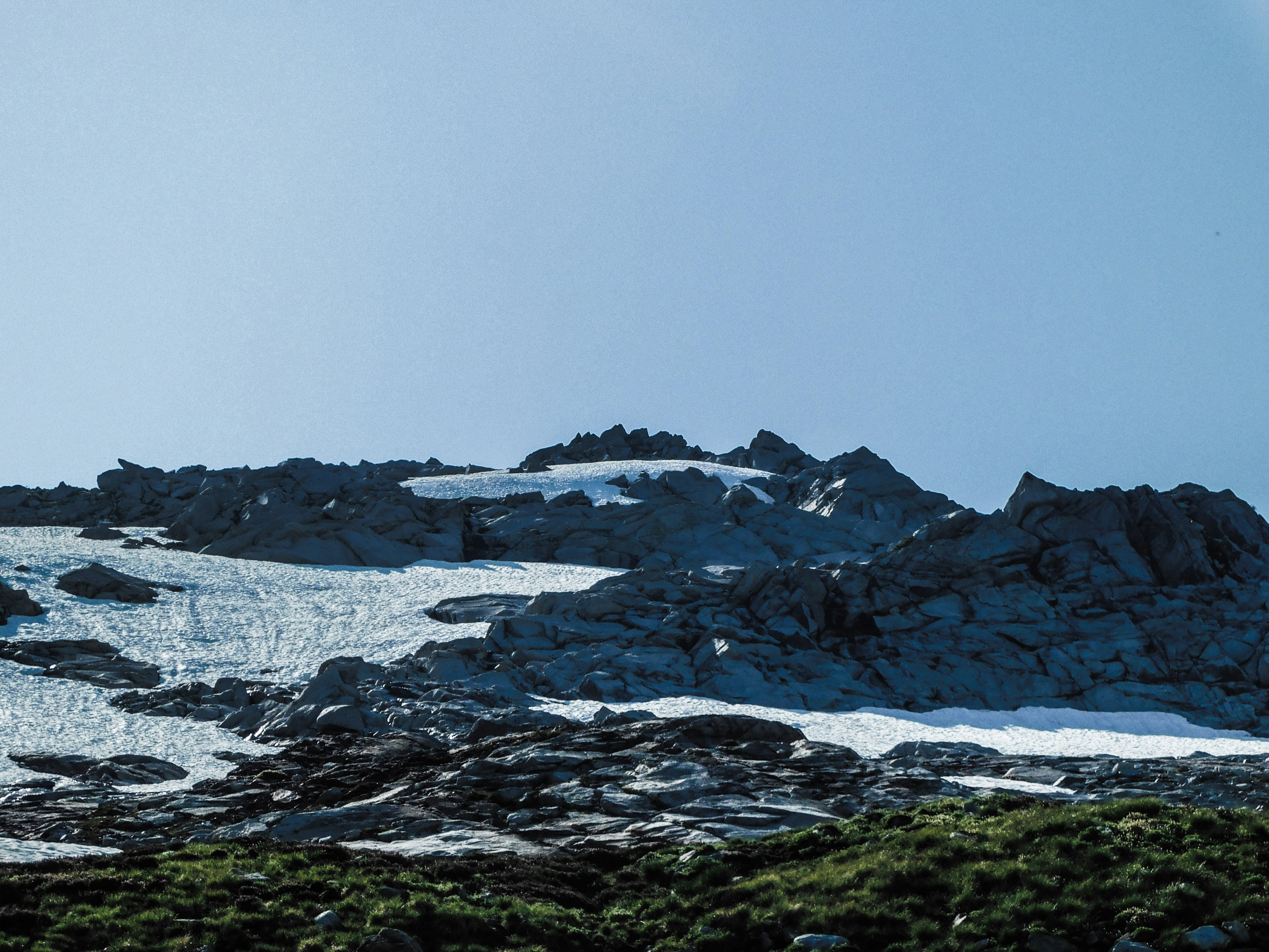 Landscape photograph of a rocky alpine ridge with snow patches and green moss, under a clear blue sky.
