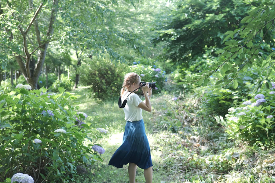 A woman stands on a leafy garden path, holding a camera up to her face amid lush greenery and blooming hydrangeas. Soft sunlight filters through the trees overhead.